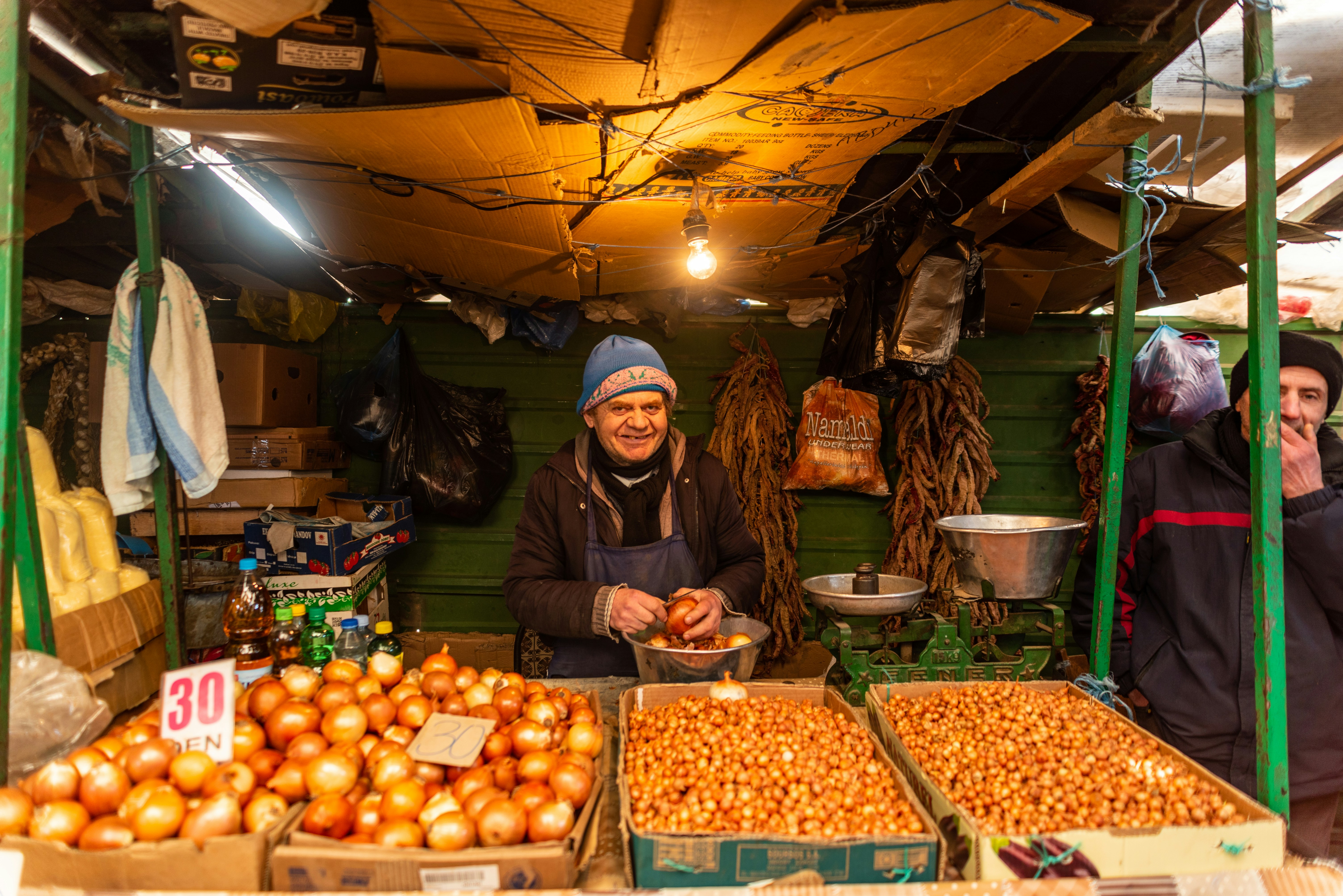 Vendor in a brown jacket stands smiling at a fruit stand filled with onions and other produce.