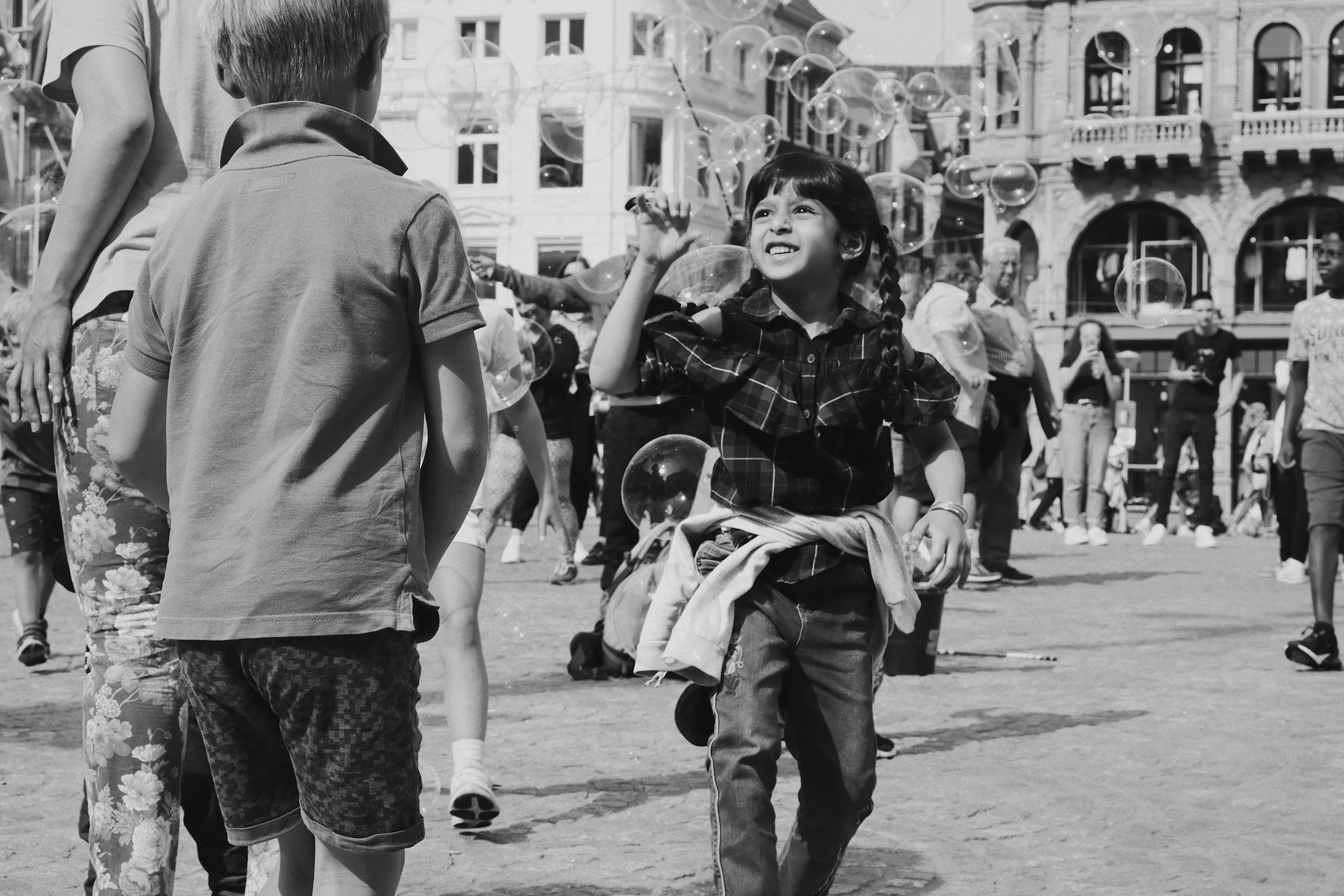 A child joyfully reaching for floating bubbles in a bustling square, surrounded by other children and adults. The scene captures a moment of pure delight and playfulness.