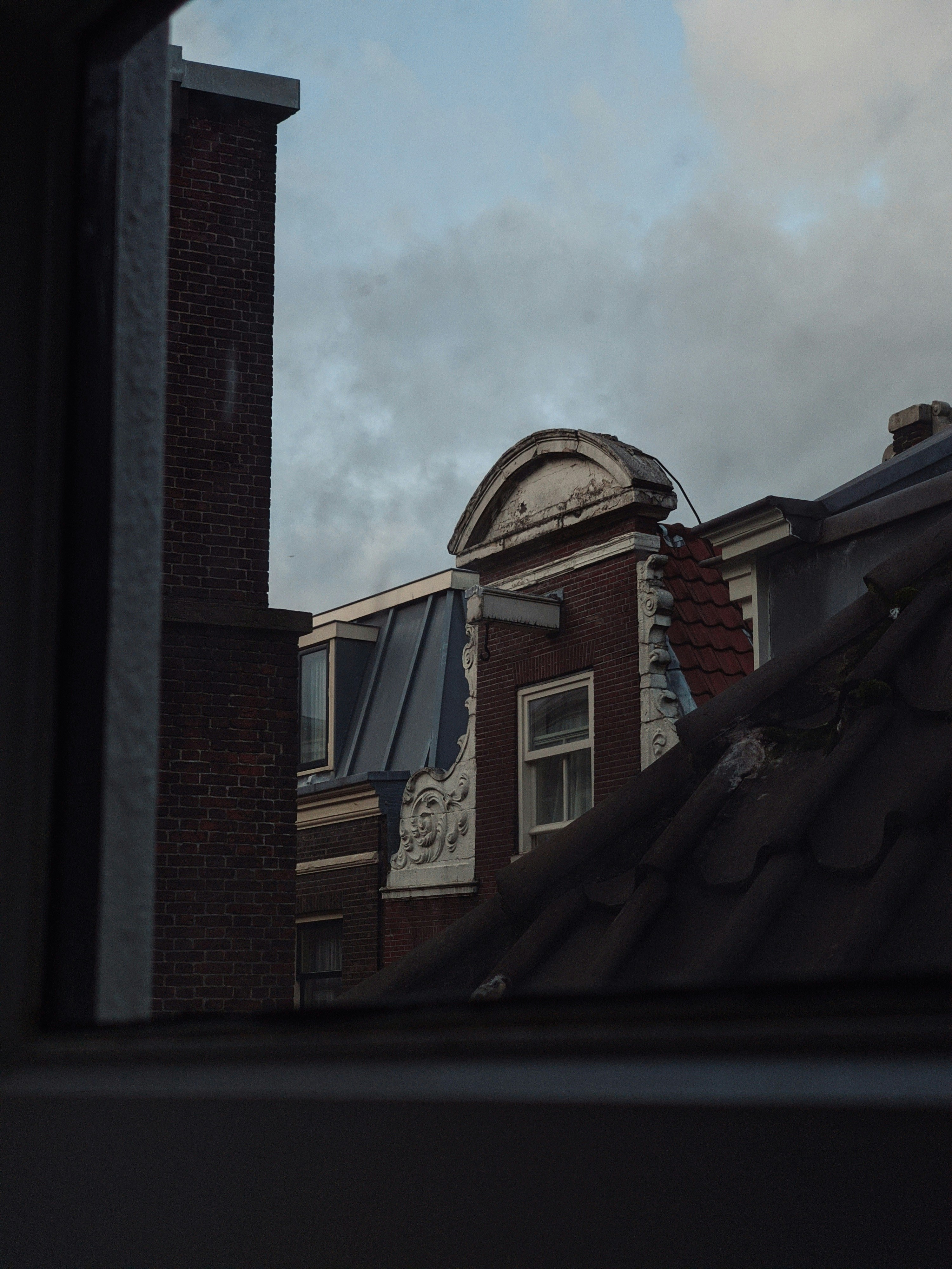 An ornate white architectural ornament crowns a red brick facade in a narrow rooftop view, framed by dark foreground eaves. The pale blue sky provides a calm backdrop.