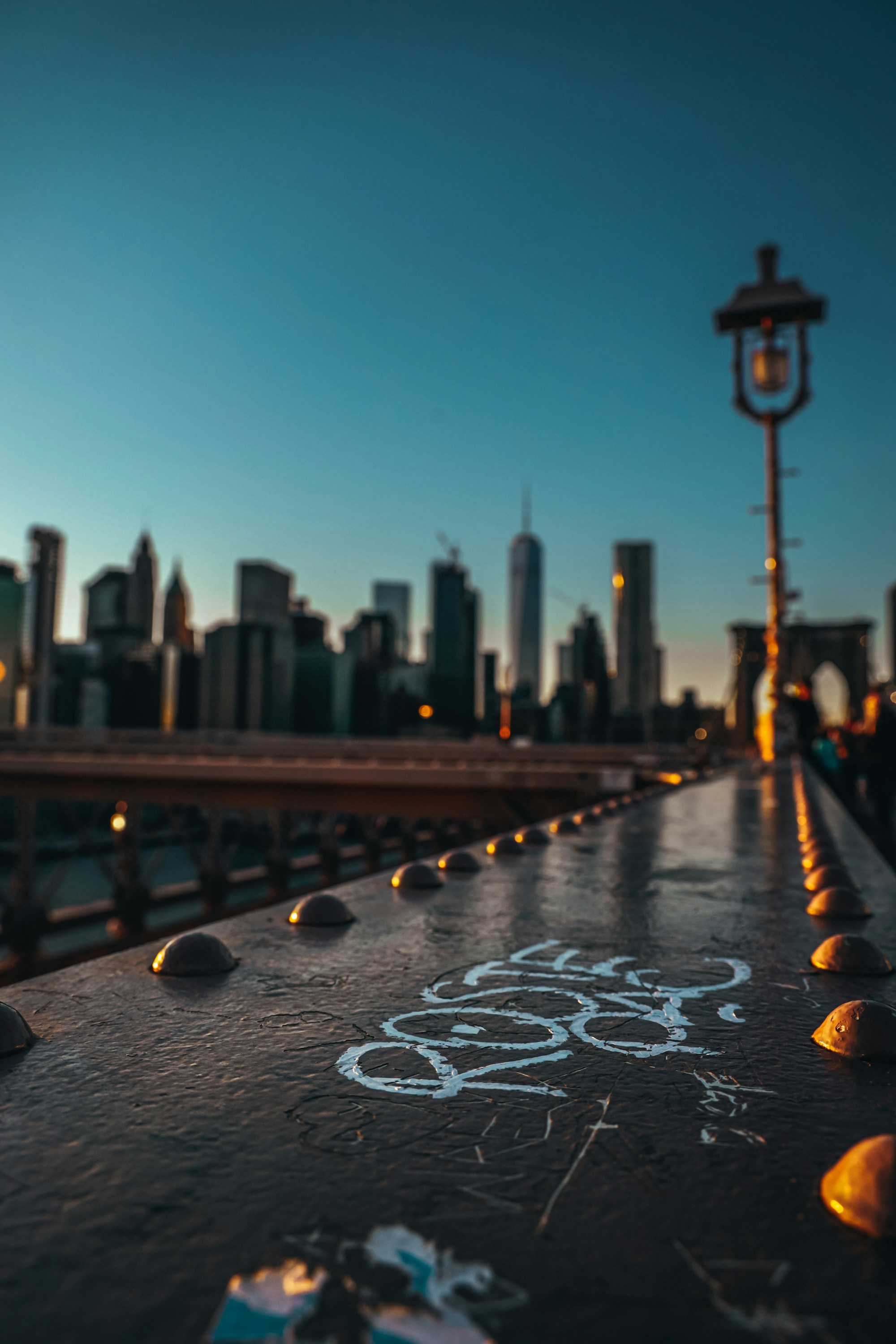 Evening light on a graffiti-marked railing with a distant city skyline and warm lamppost glow. A moody, urban bridge scene captured with shallow depth of field.