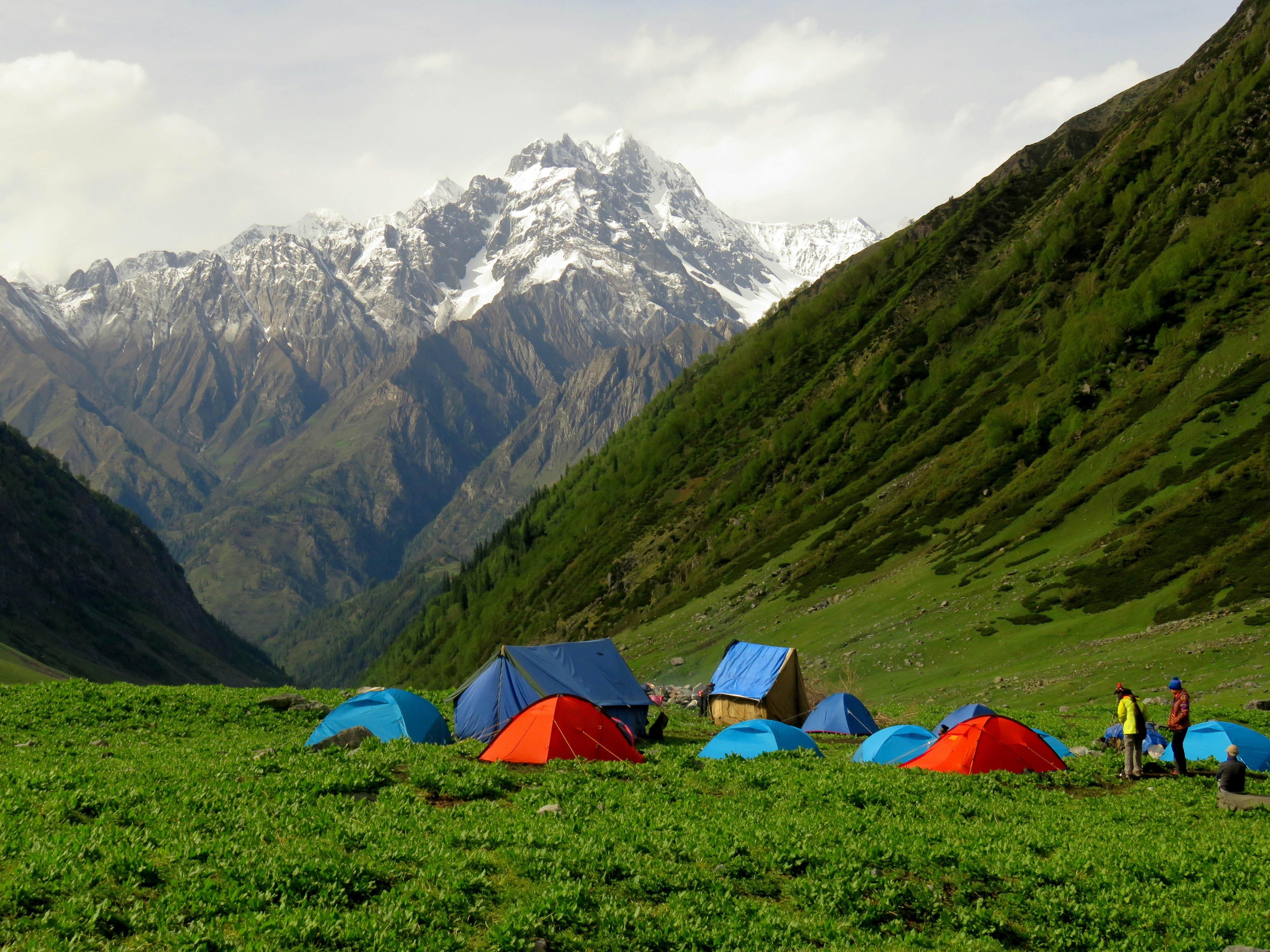 Colorful tents nestled in a lush green valley surrounded by towering snow-capped mountains. The scene captures the essence of outdoor adventure and tranquility.