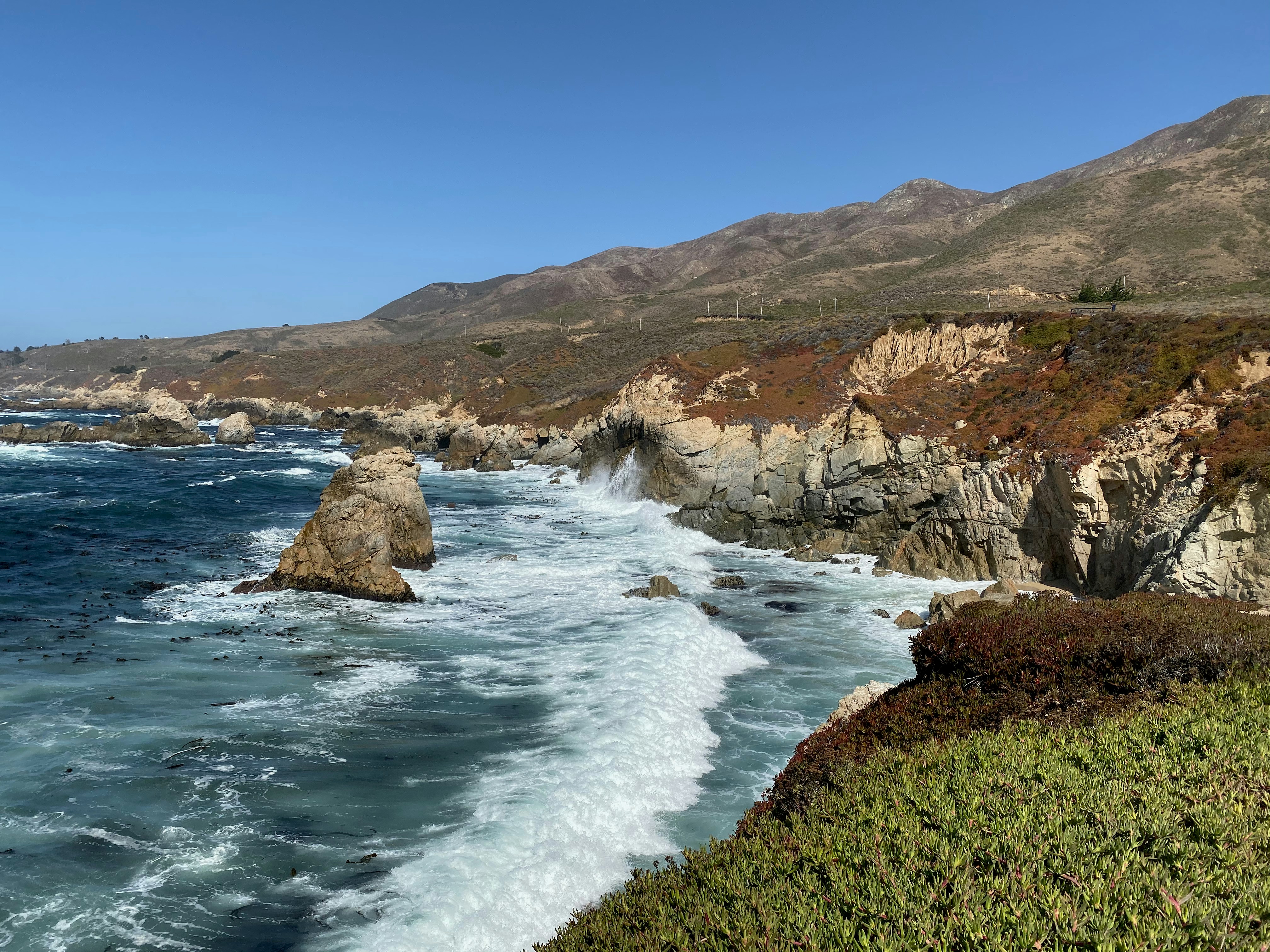Waves crash against rugged cliffs under a clear blue sky, showcasing the dynamic interaction between land and ocean.