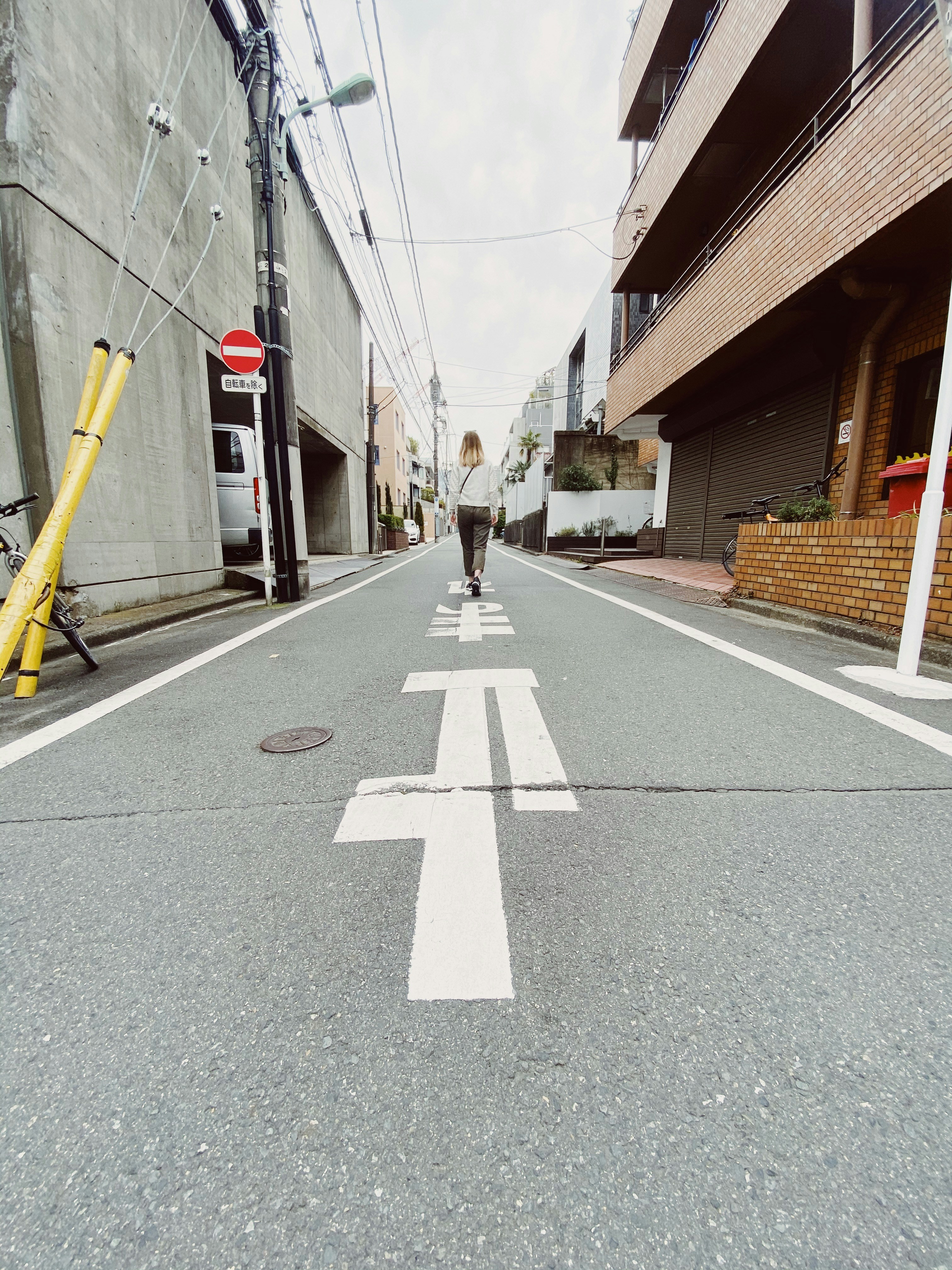 A person walking down a narrow urban street, flanked by modern buildings and utility poles, leading to an unseen destination.