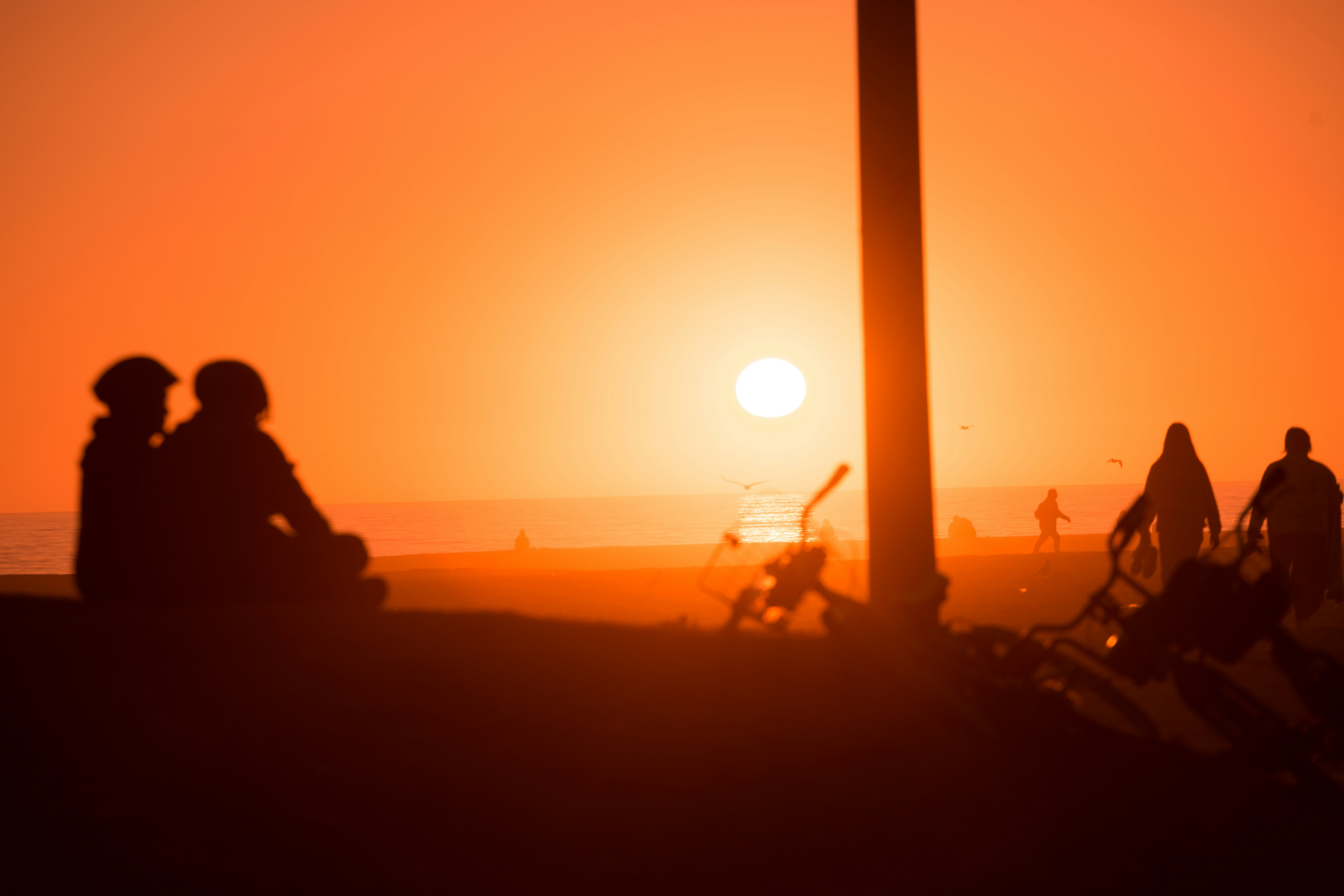 Silhouetted figures enjoying a sunset at the beach, with bicycles resting nearby. The sun casts a warm glow over the scene.