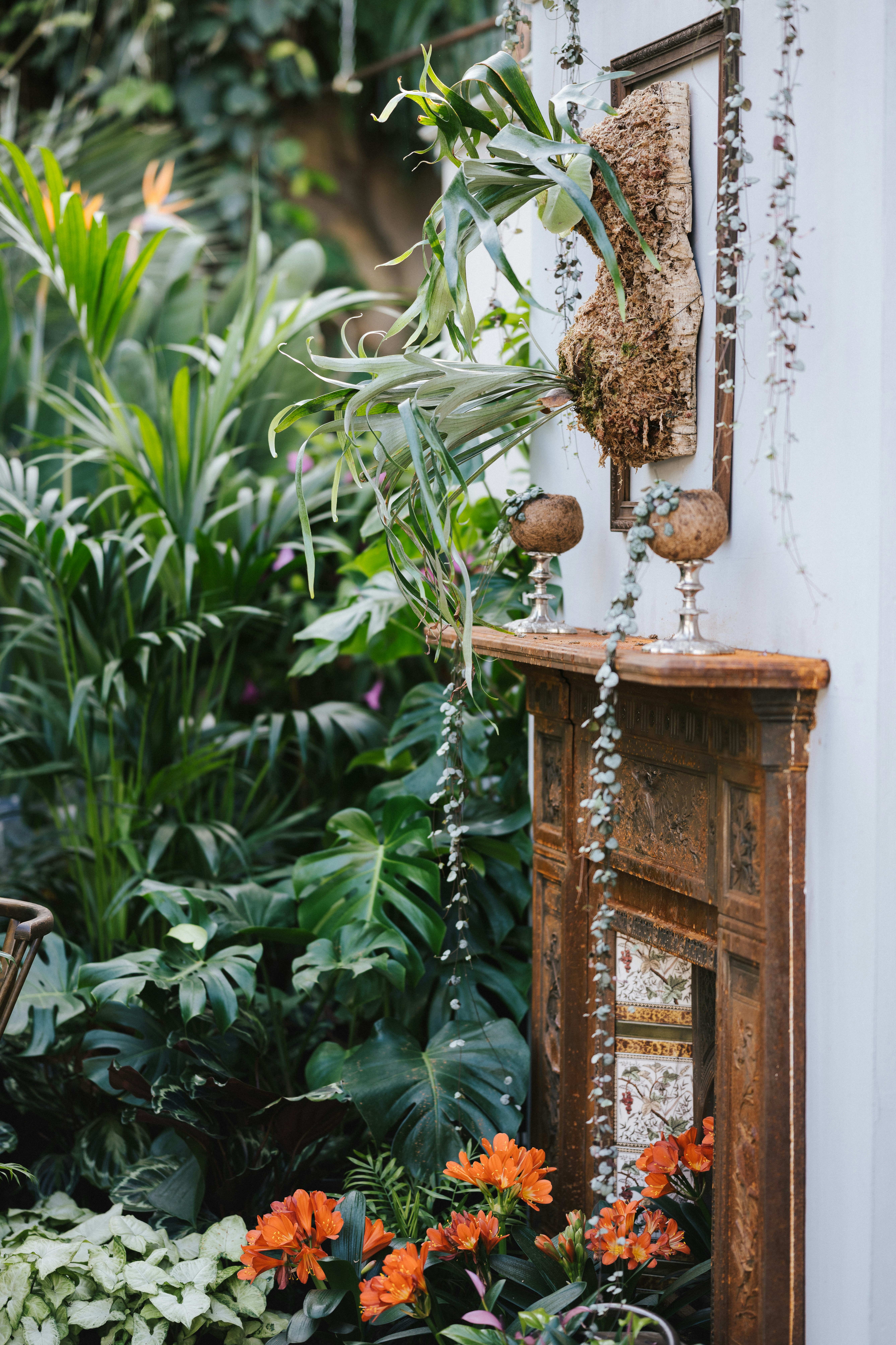 Lush greenery surrounds an ornate fireplace adorned with vibrant orange flowers and a unique plant display, creating a tranquil indoor garden atmosphere.