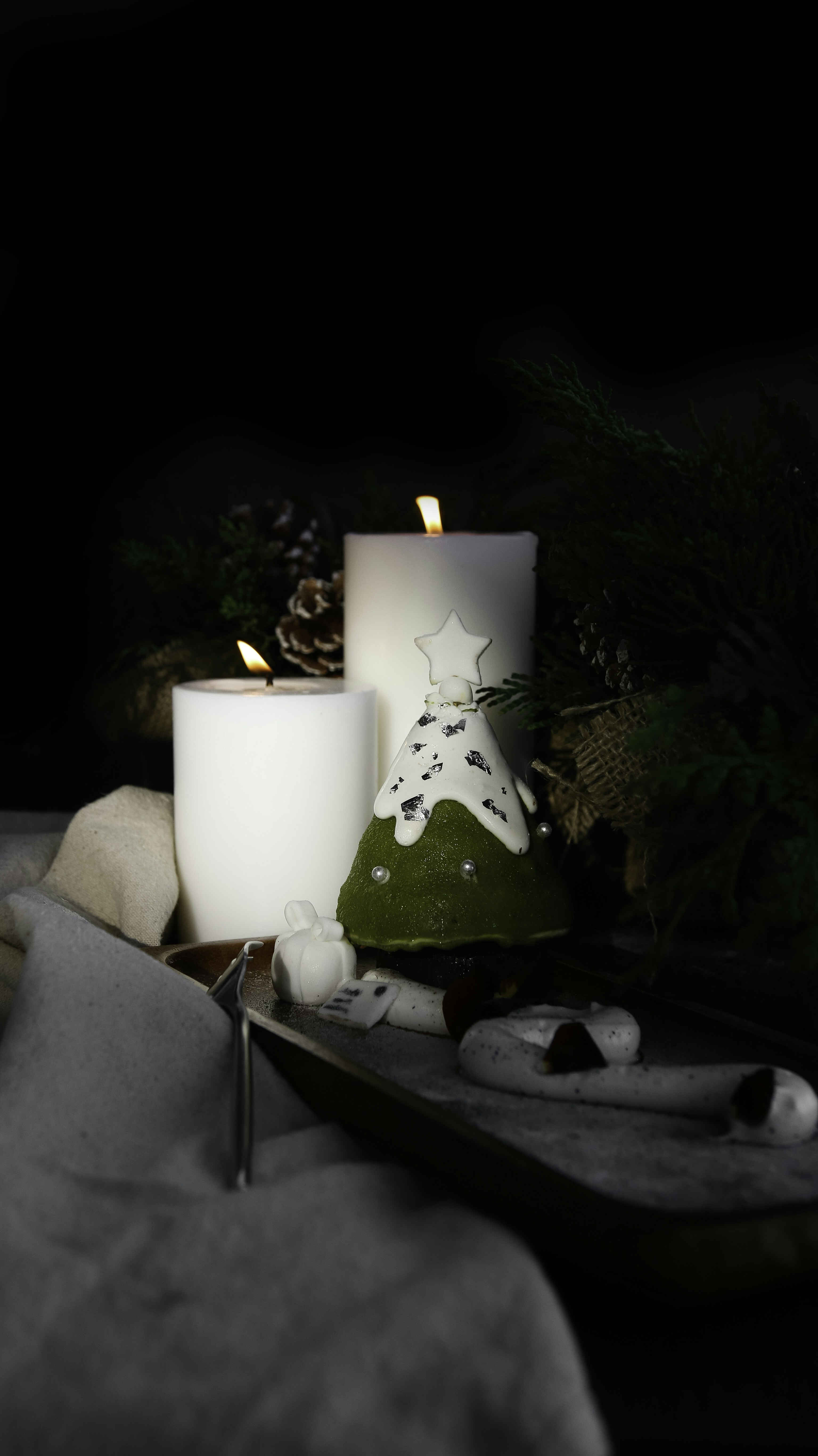 A green Christmas tree-shaped dessert adorned with white icing sits on a wooden platter beside glowing candles and pinecones, evoking a cozy holiday atmosphere.