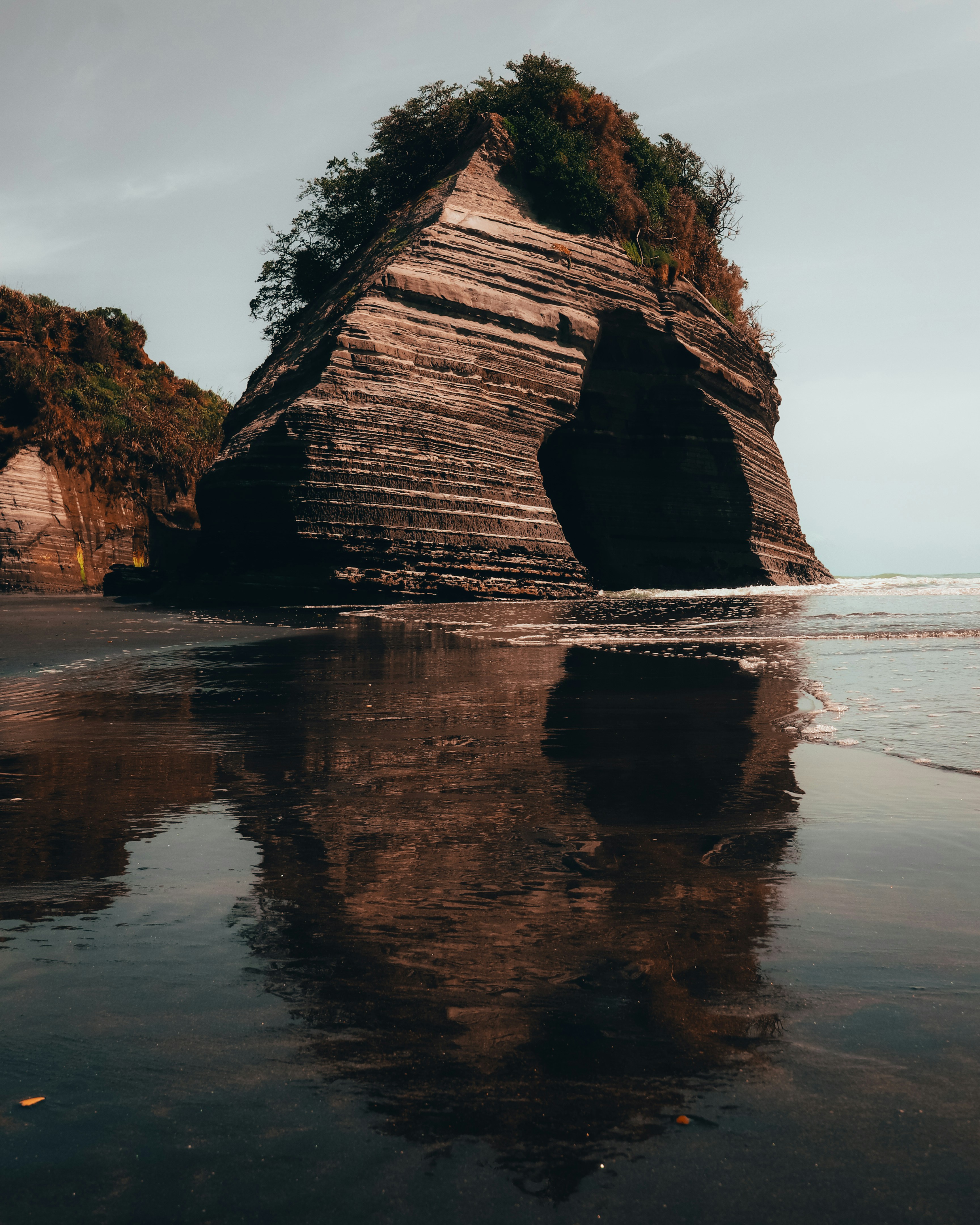 A dramatic rock formation rises from the shoreline, mirrored in the wet sand below, surrounded by lush greenery. The scene captures the raw beauty of coastal landscapes.