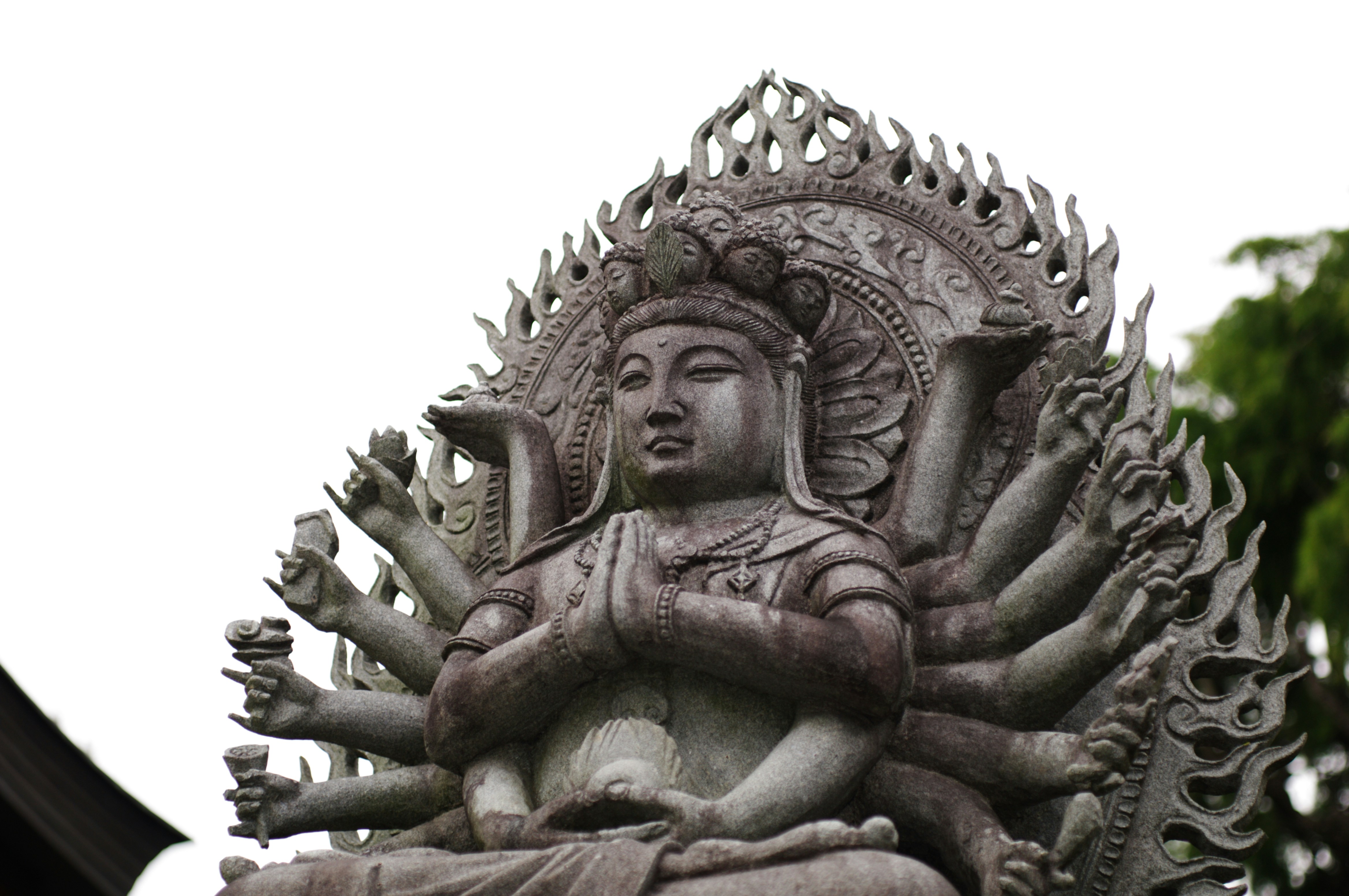 Stone sculpture of Senju-Kannon with multiple arms, set against a bright sky and greenery.