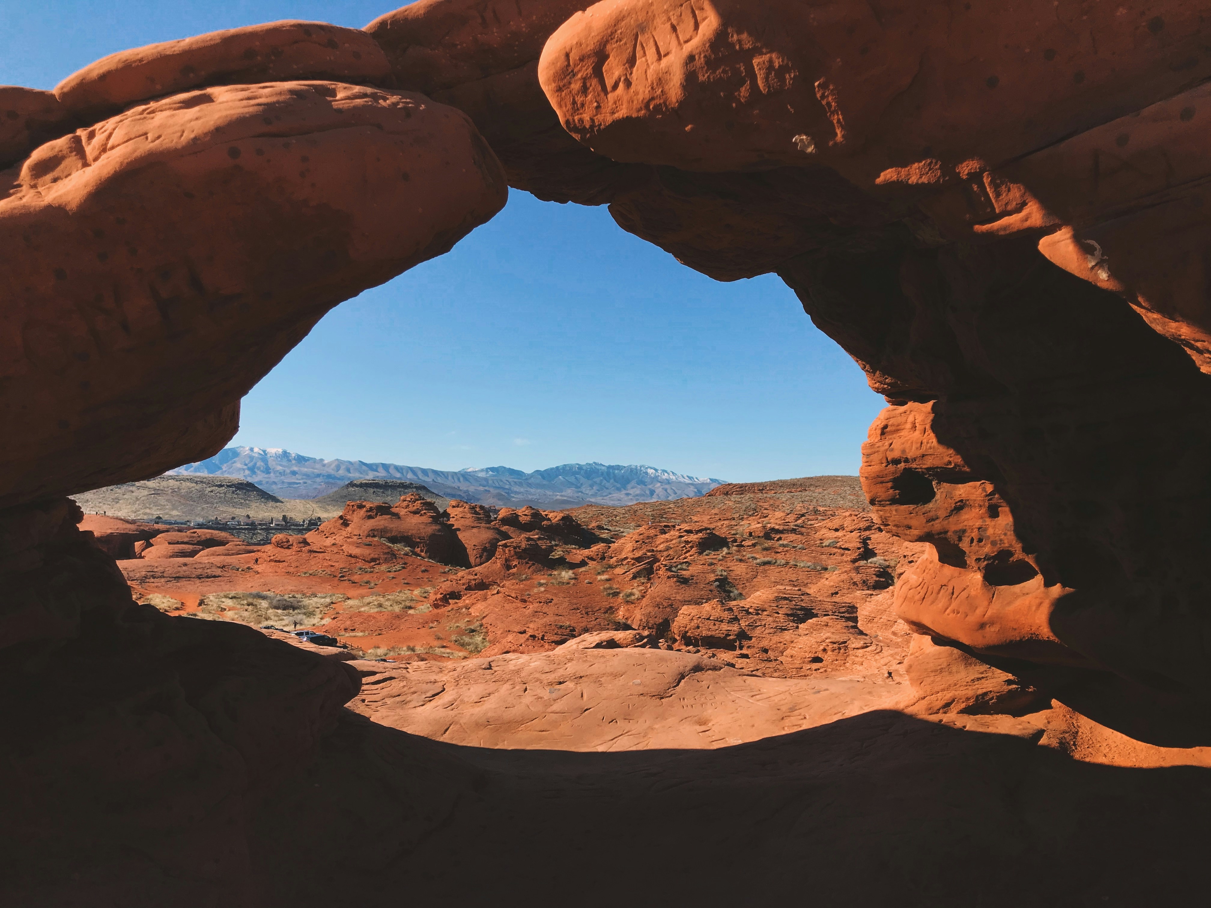 View through a natural rock arch, showcasing vibrant red rock formations and distant mountains under a clear blue sky.