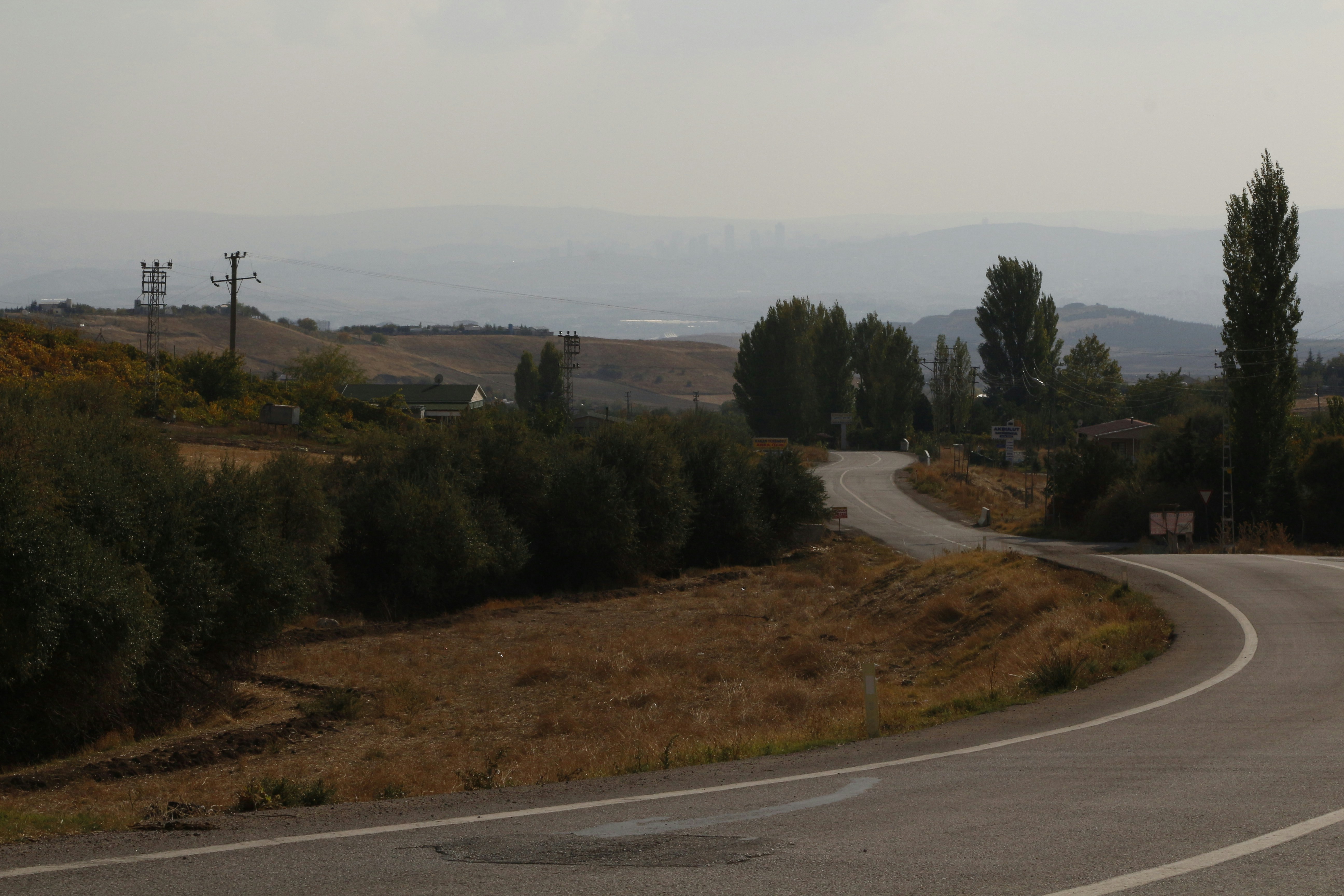 Curving road meanders through a tranquil countryside, framed by gentle hills and distant mountains under a hazy sky.