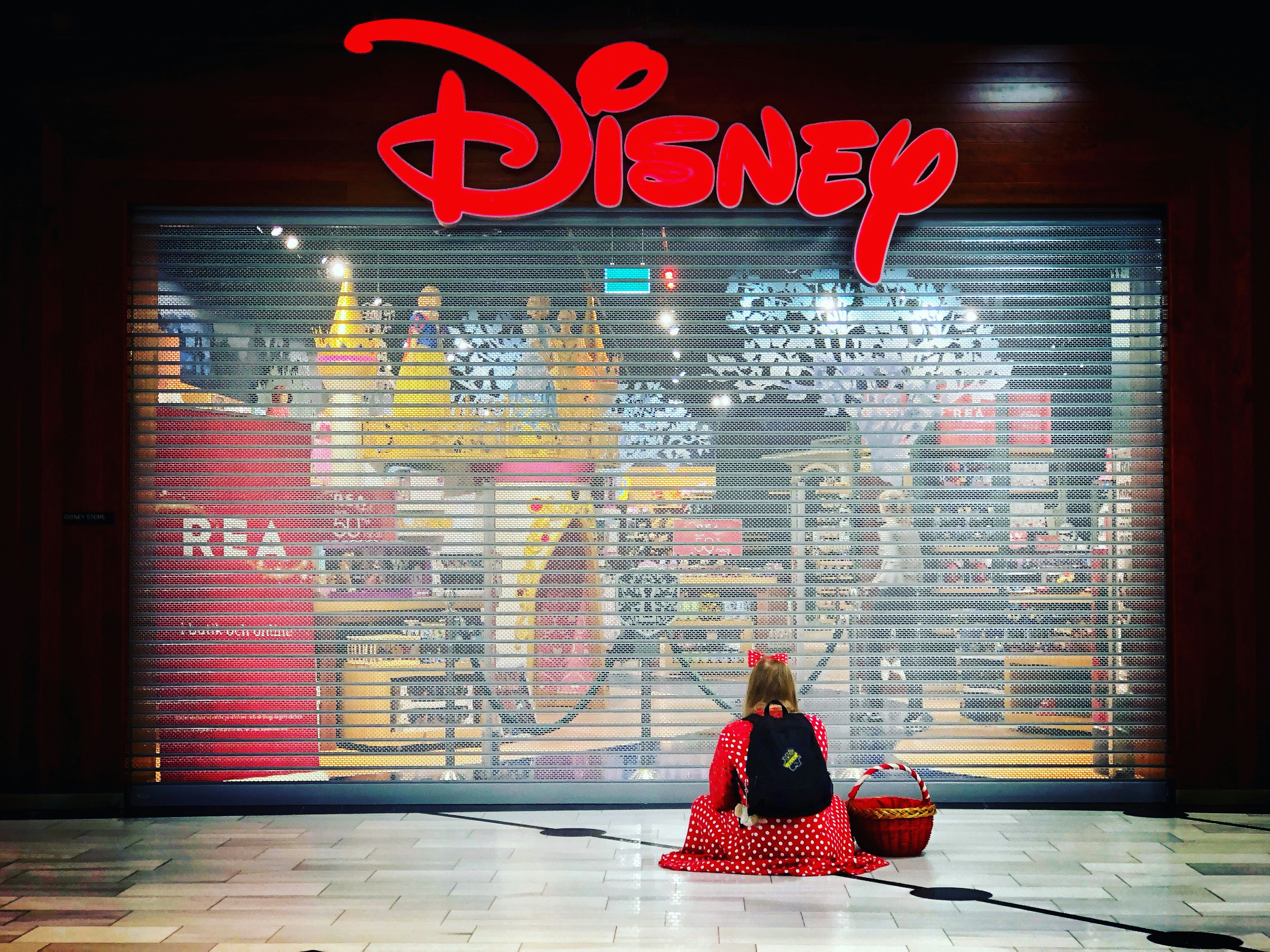 Person in red and black attire sitting in front of a closed Disney store with a reflective metal shutter.