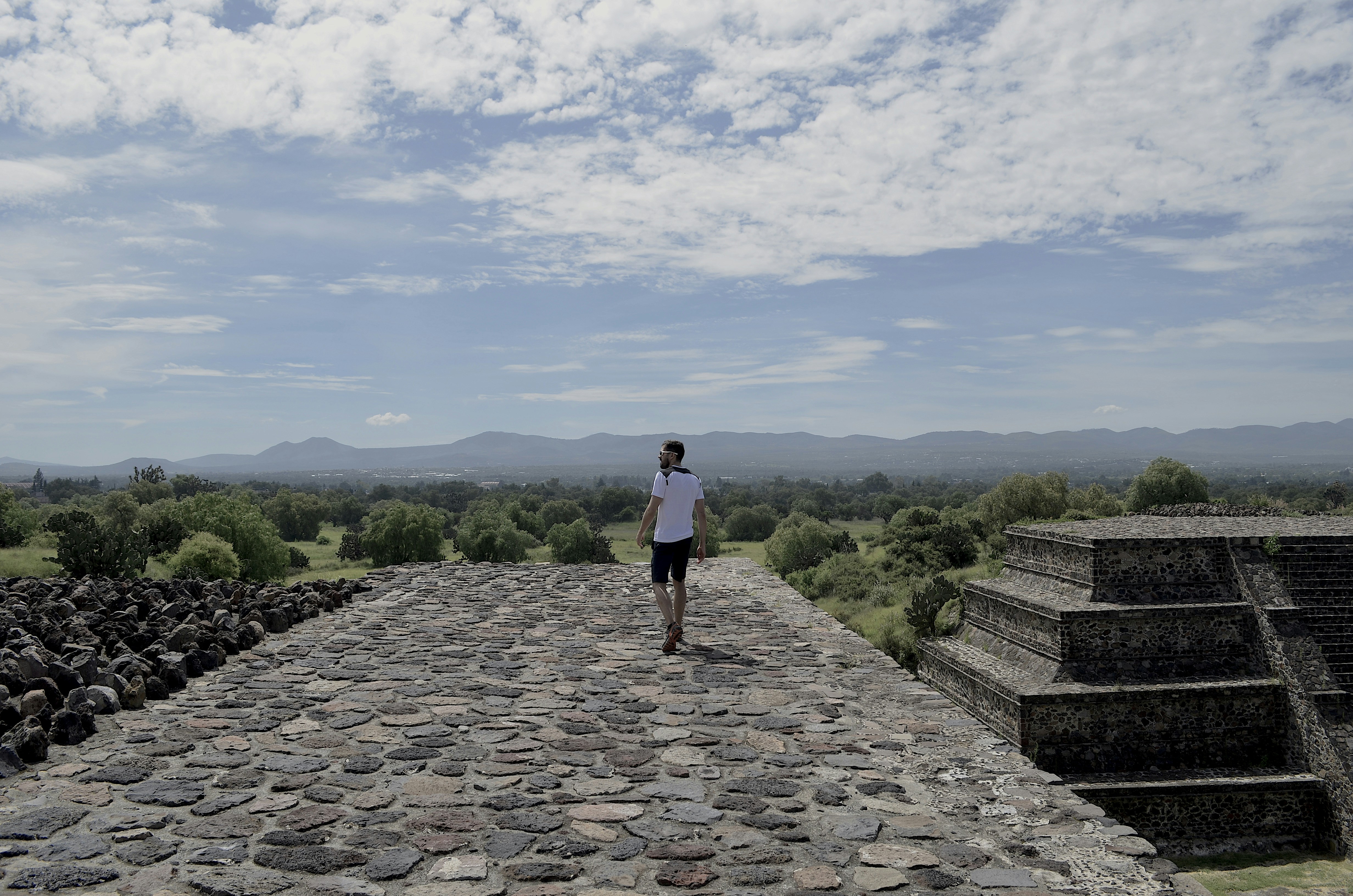 Person walking on a stone pathway surrounded by historical ruins and vast landscape under a cloudy sky.