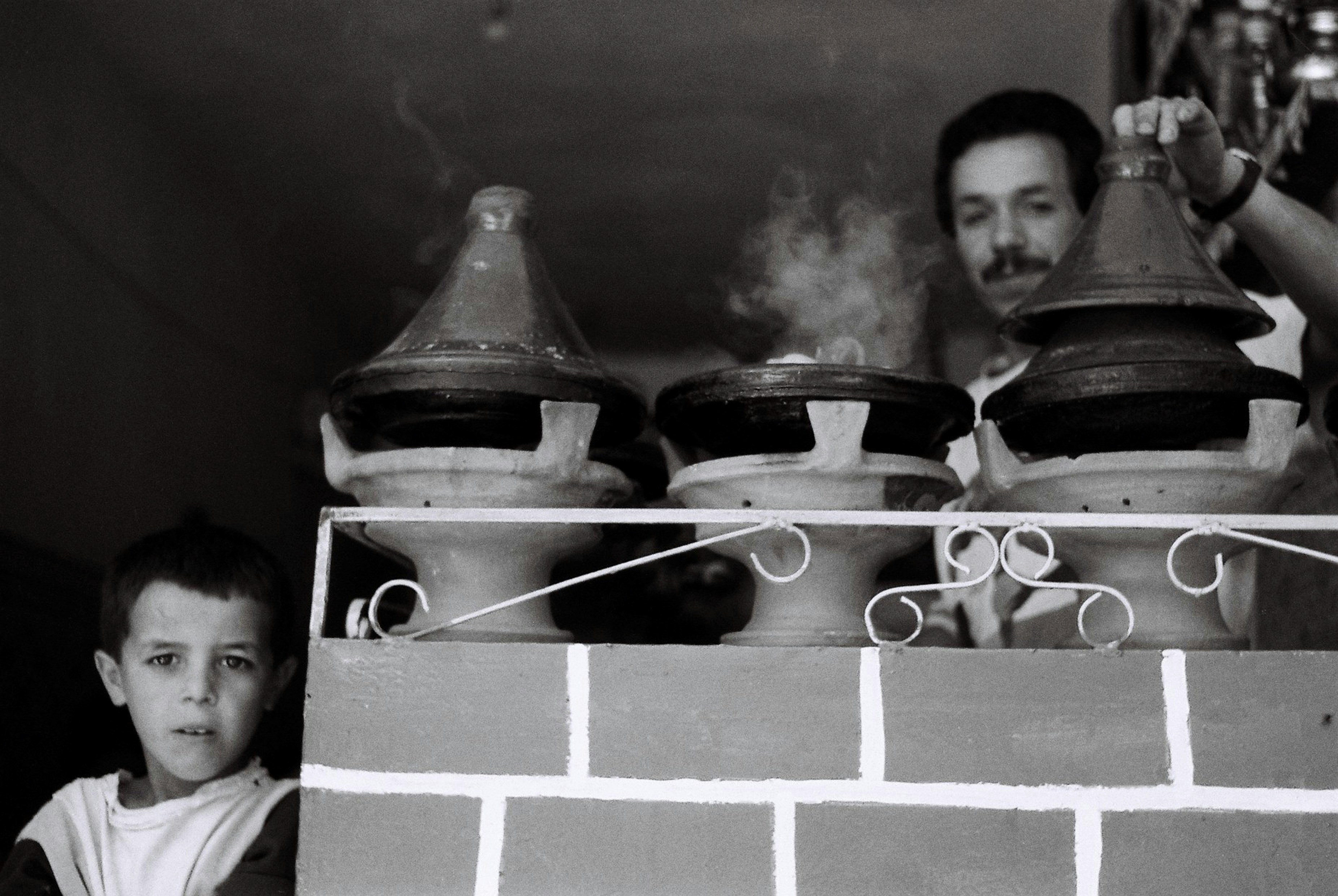 A young boy watches intently as a man prepares traditional clay pots, releasing wisps of steam in a rustic kitchen setting.