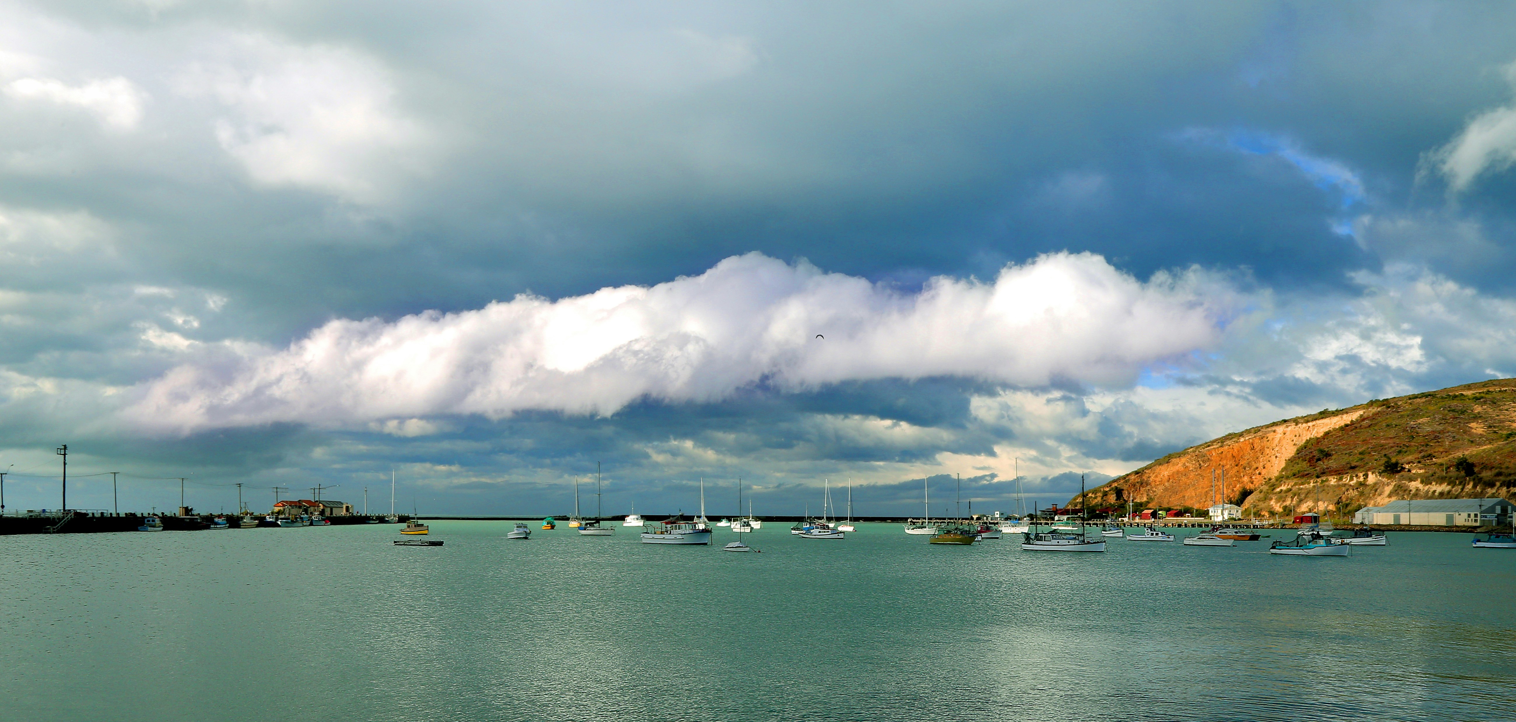 Fishing boats rest in a serene harbor beneath dramatic cloud formations and a rugged hillside.