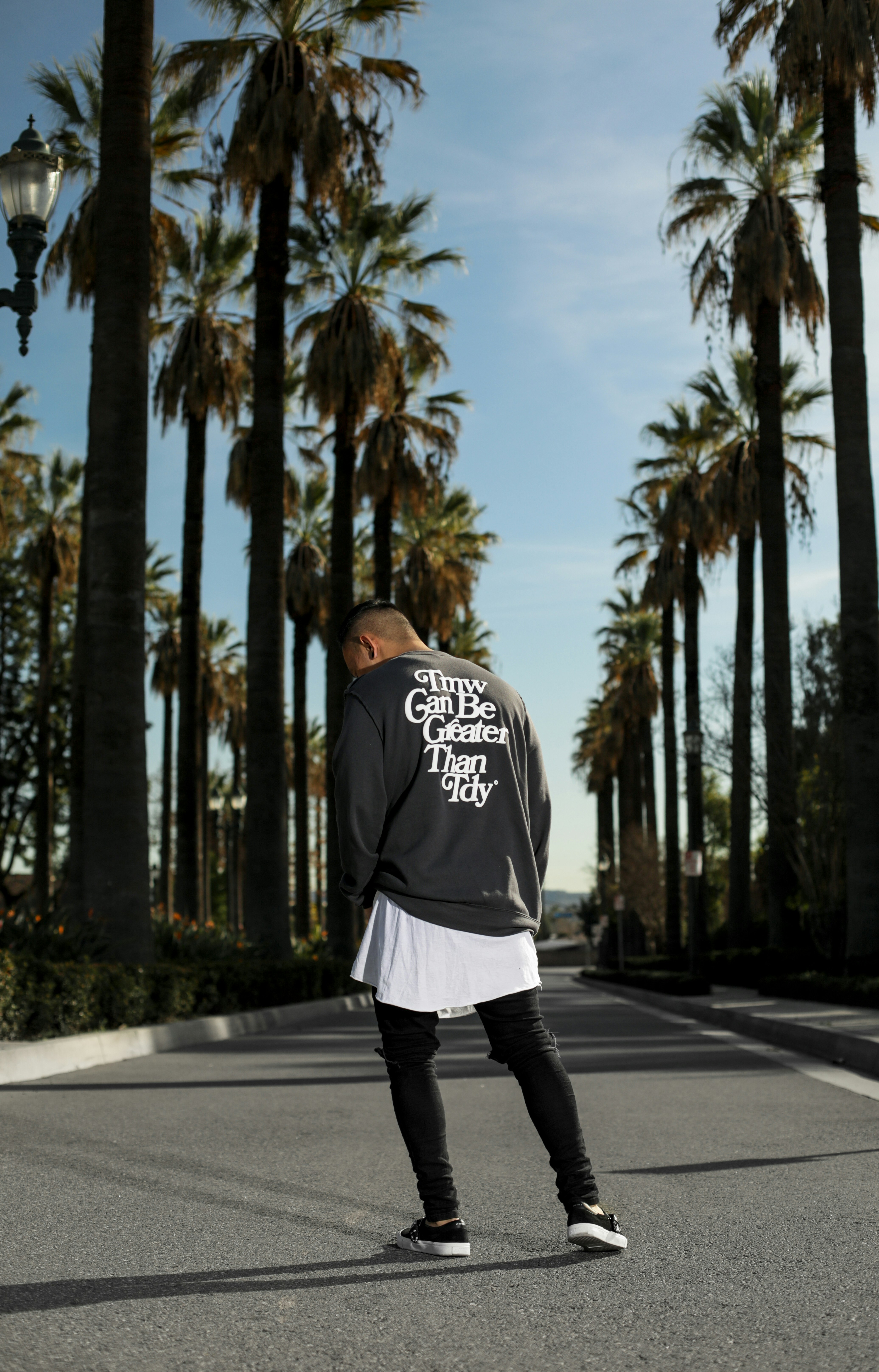 Person standing on a palm tree-lined street, wearing a shirt with a motivational message under a clear blue sky.