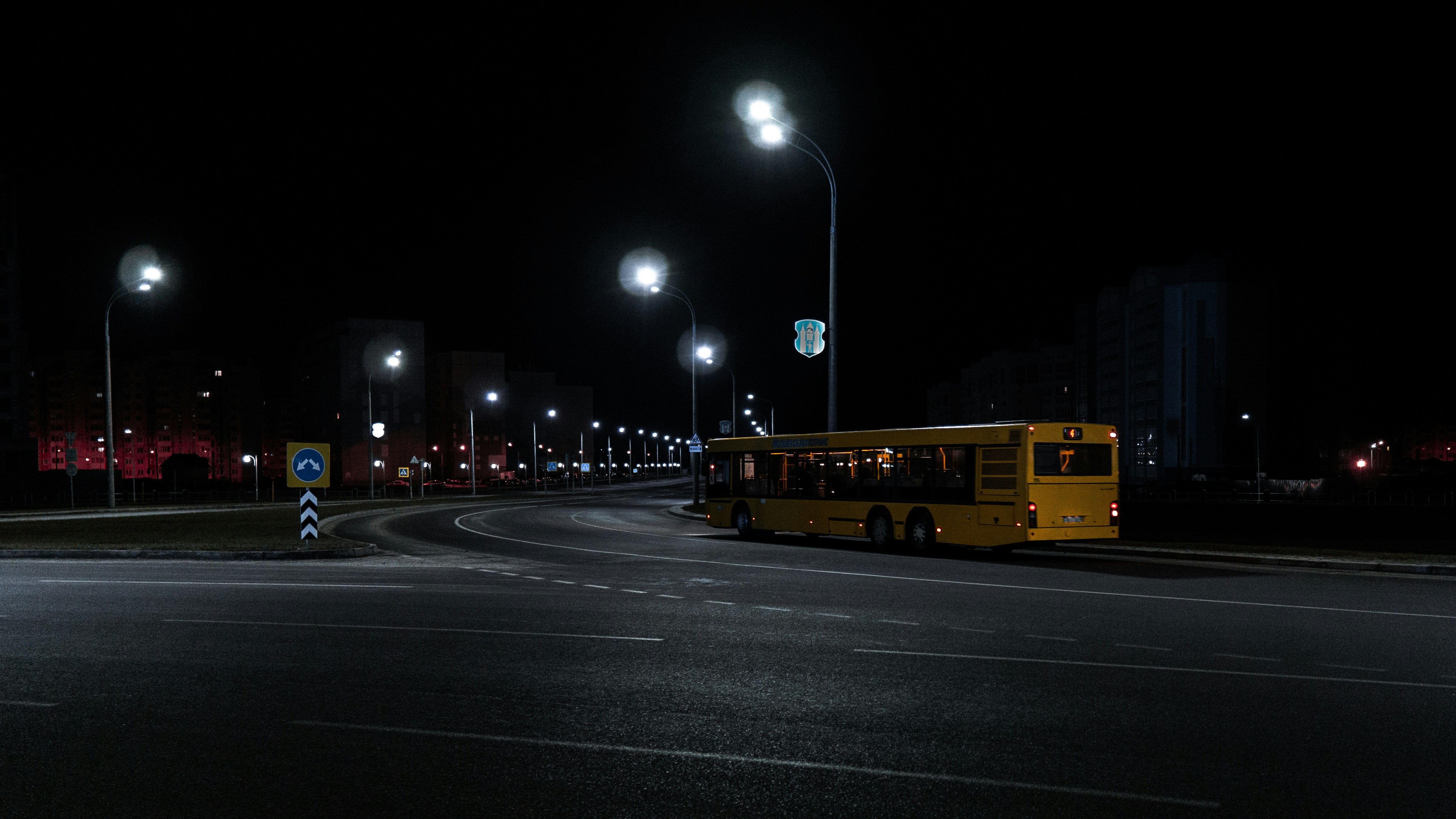 A yellow bus waits at a dimly lit roundabout, surrounded by urban architecture and streetlights illuminating the night. 