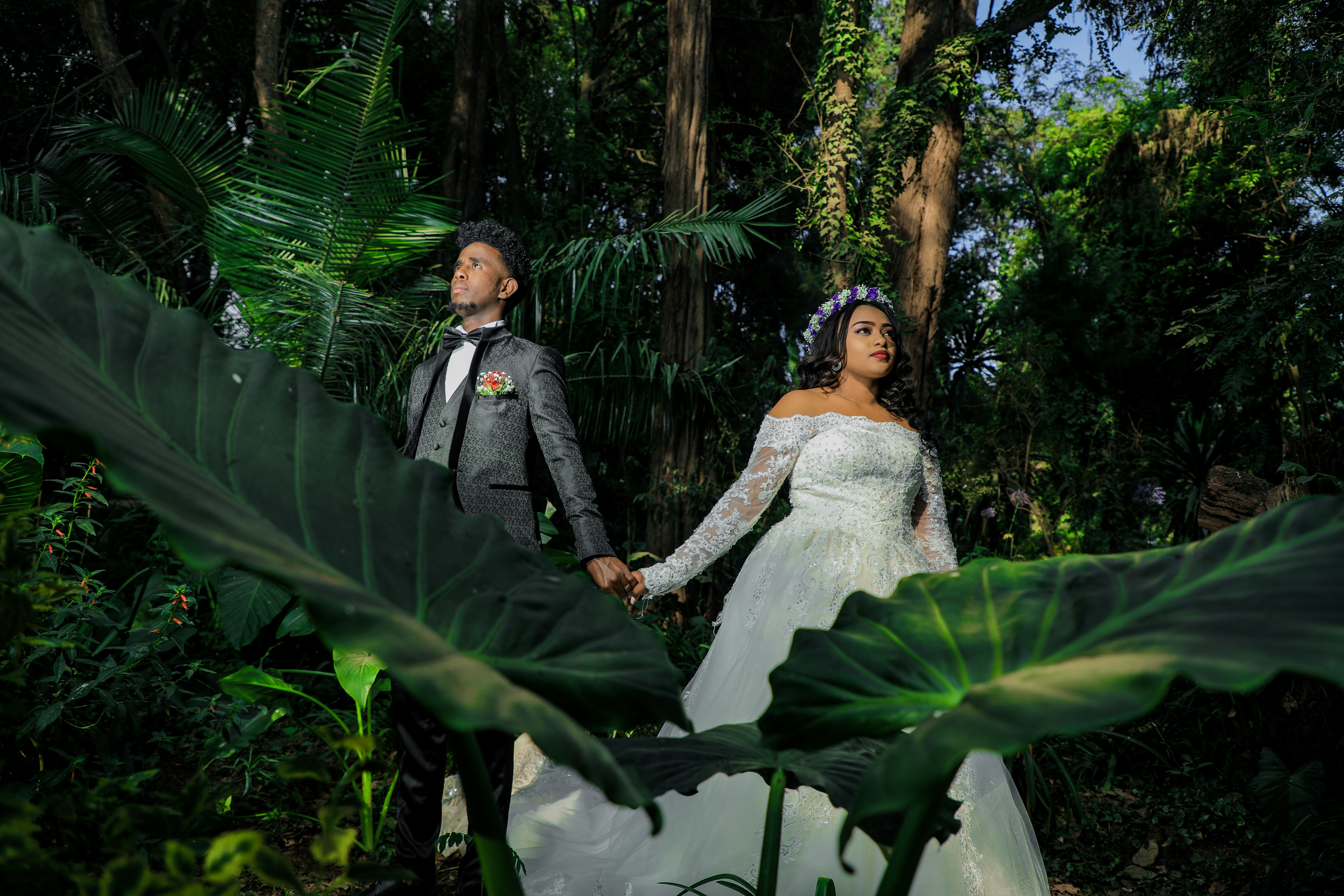 Couple holding hands in a verdant setting, surrounded by large tropical leaves, showcasing a moment of unity. The bride's elegant gown contrasts beautifully with the natural surroundings.