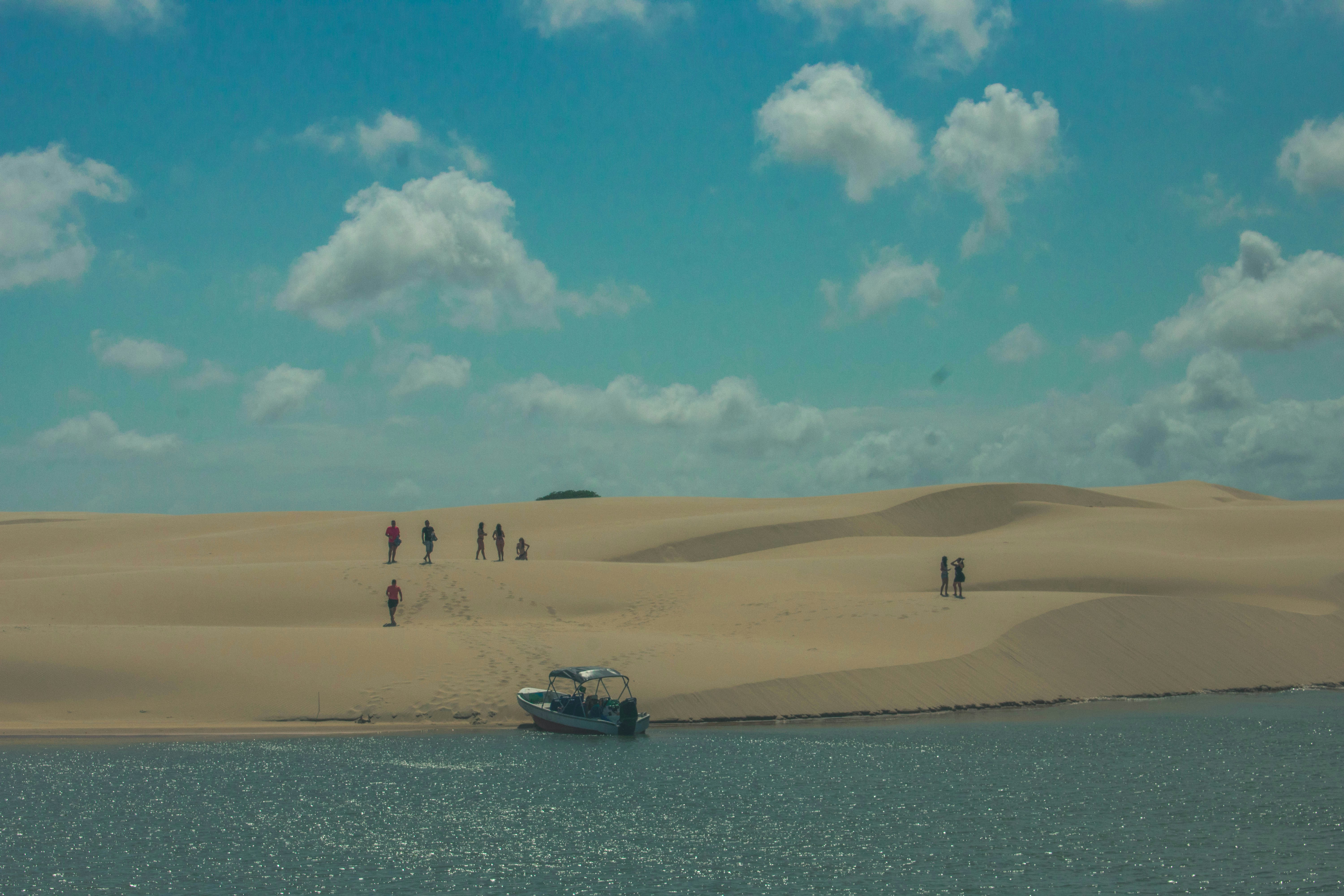 People walking across sandy dunes under a partly cloudy sky with a boat moored by the water's edge.