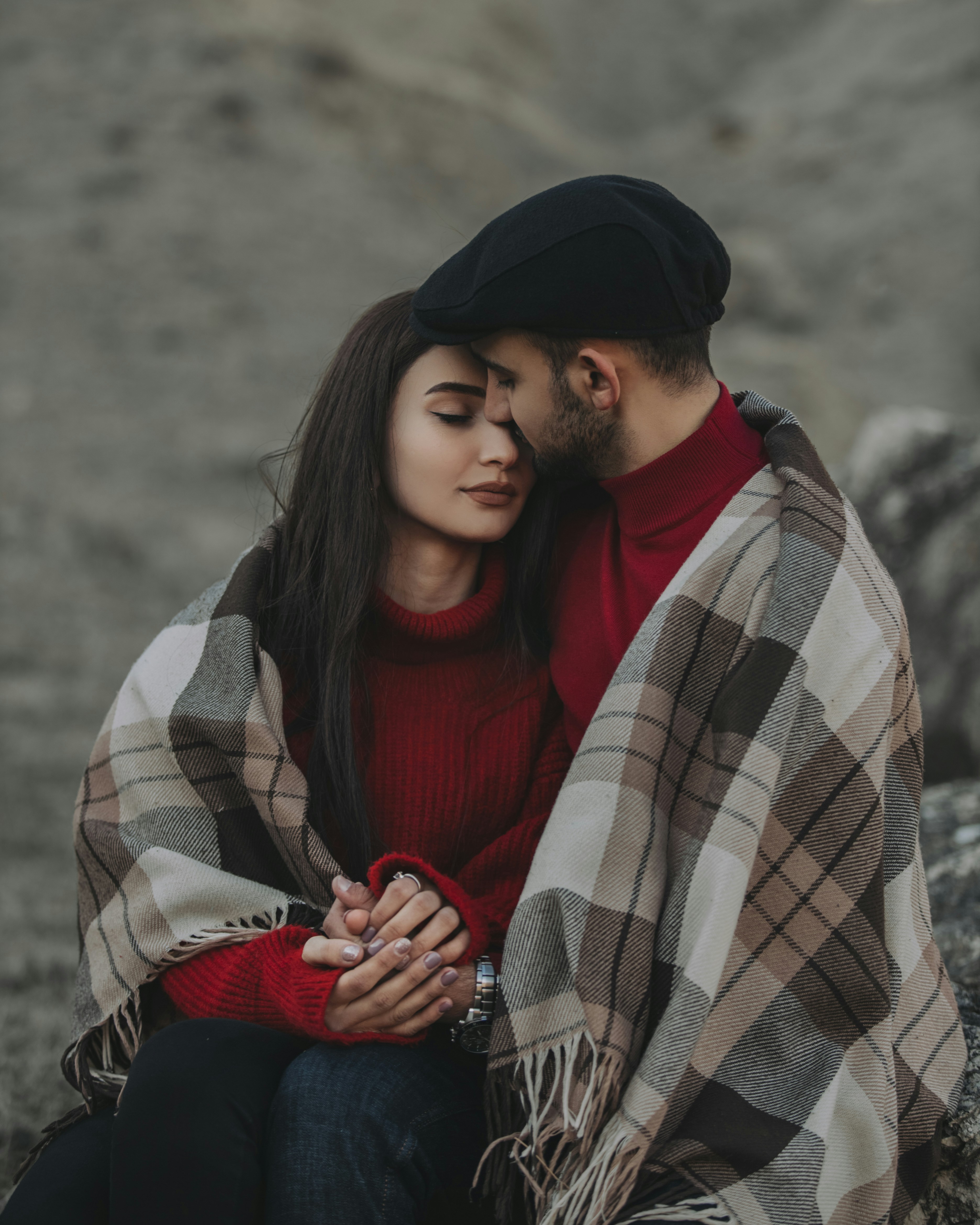 Couple wrapped in a plaid blanket, sharing an intimate moment on a rocky landscape, dressed in cozy red attire.