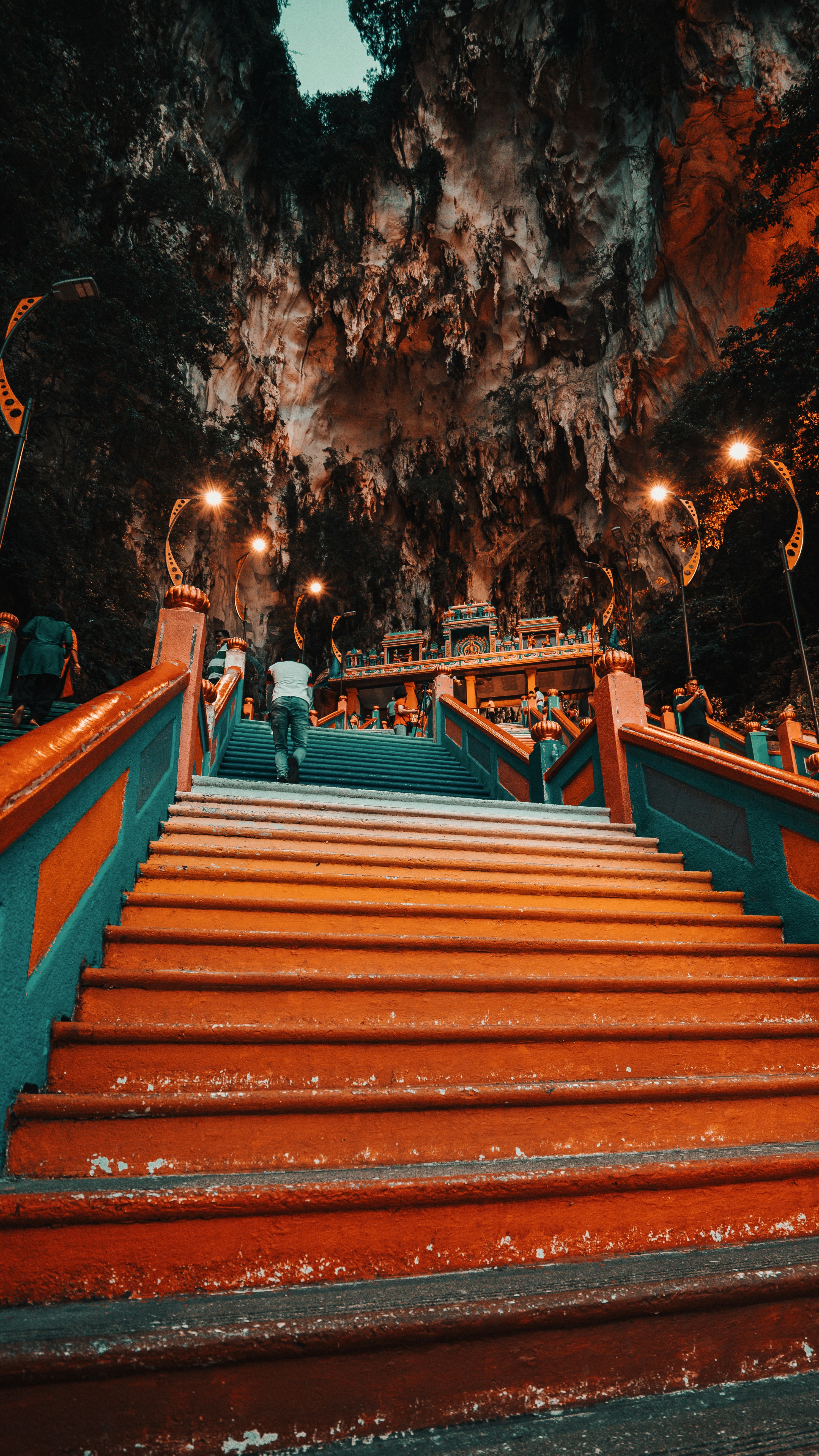 Vibrant staircase leading up to Batu Caves, framed by towering limestone cliffs and illuminated by warm lights. Visitors ascend towards the temple nestled within the cave.