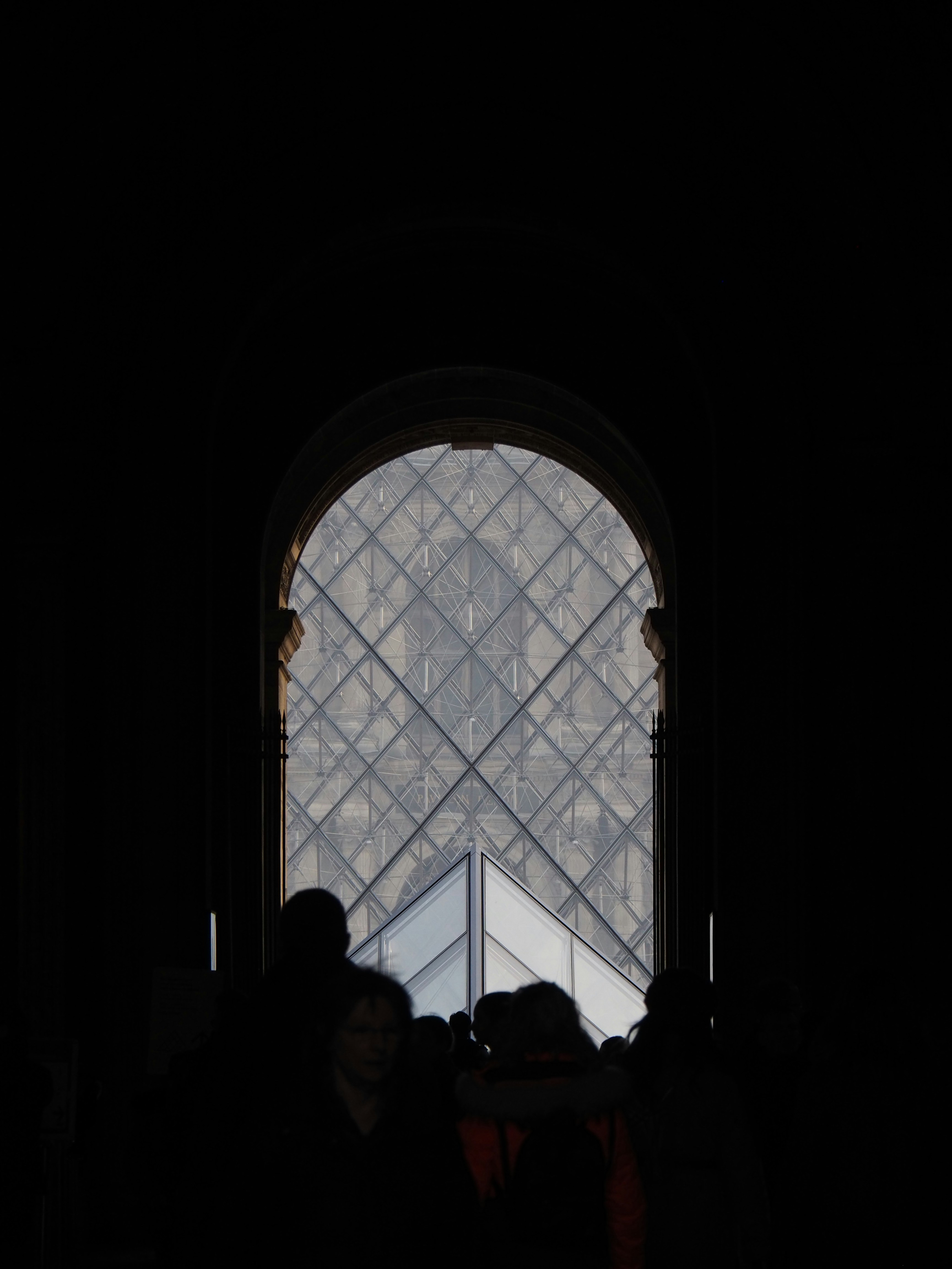 Silhouetted figures move through an archway, revealing the intricate glass pyramid of the Louvre in the background.