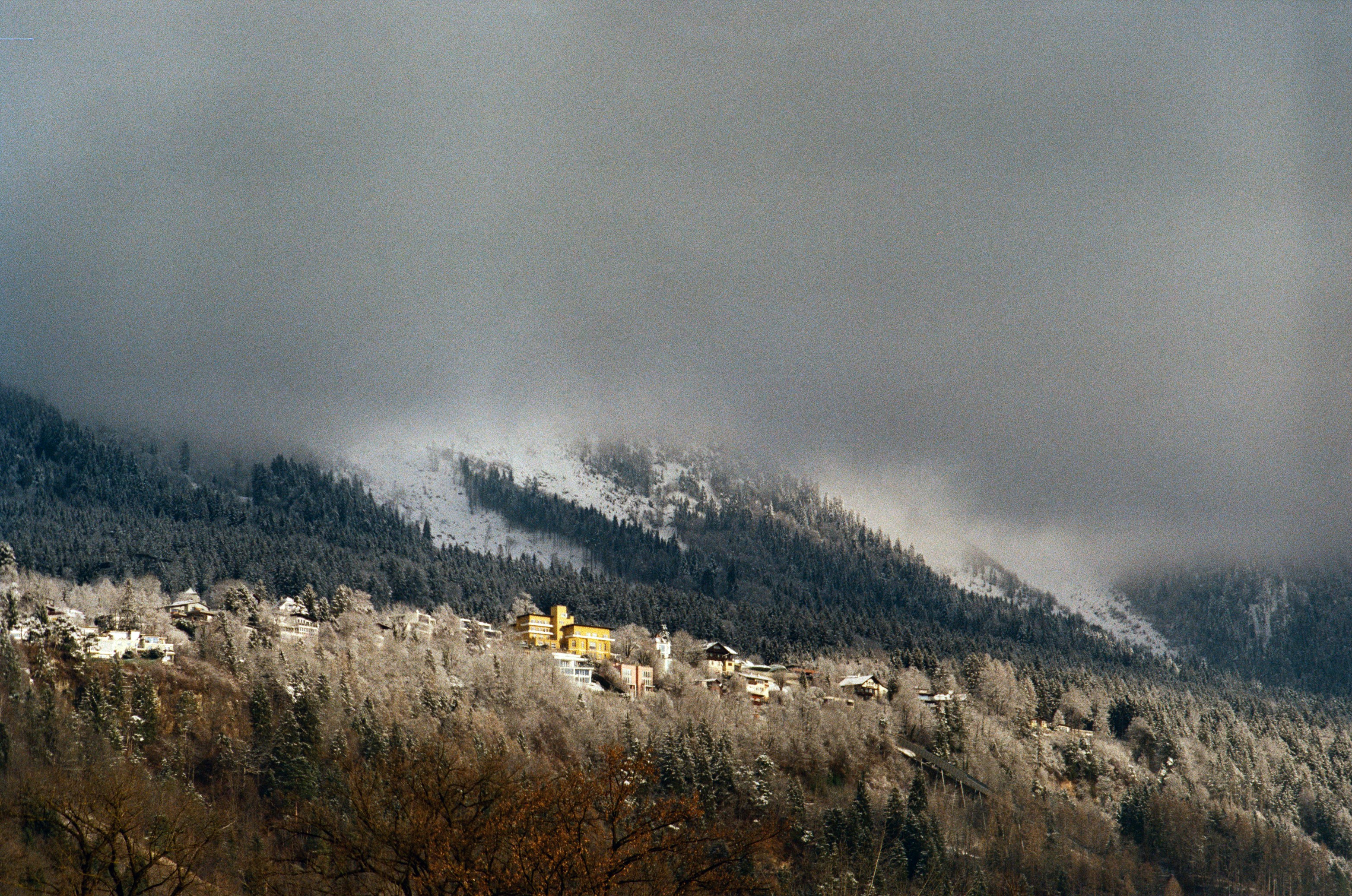 Snow-dusted hillside village on a forested slope as heavy clouds drift over alpine peaks. A quiet winter landscape captured in a photograph.