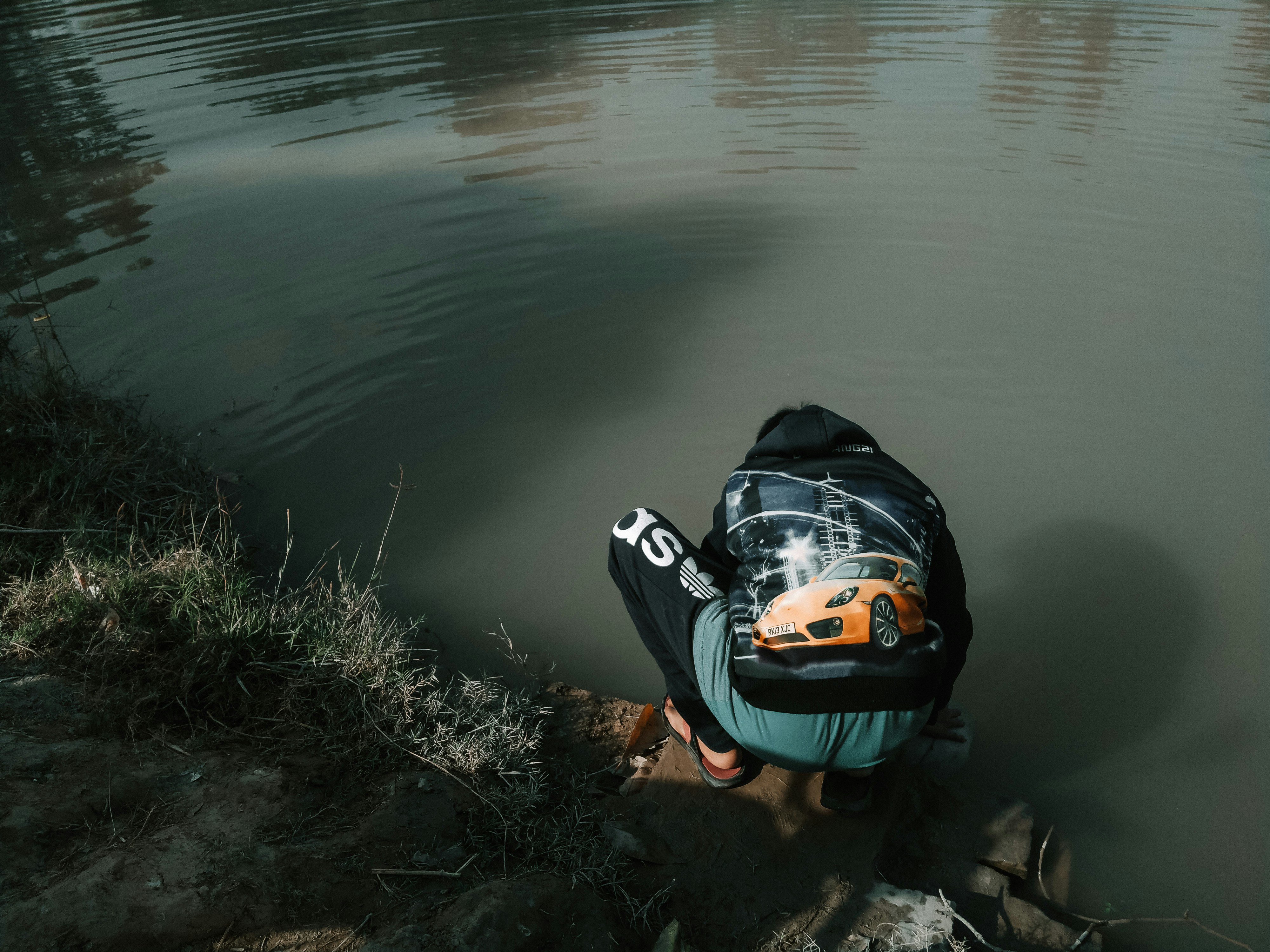 Individual crouching near the water, examining the surface with a playful car design on their back. The scene captures a moment of exploration and wonder.