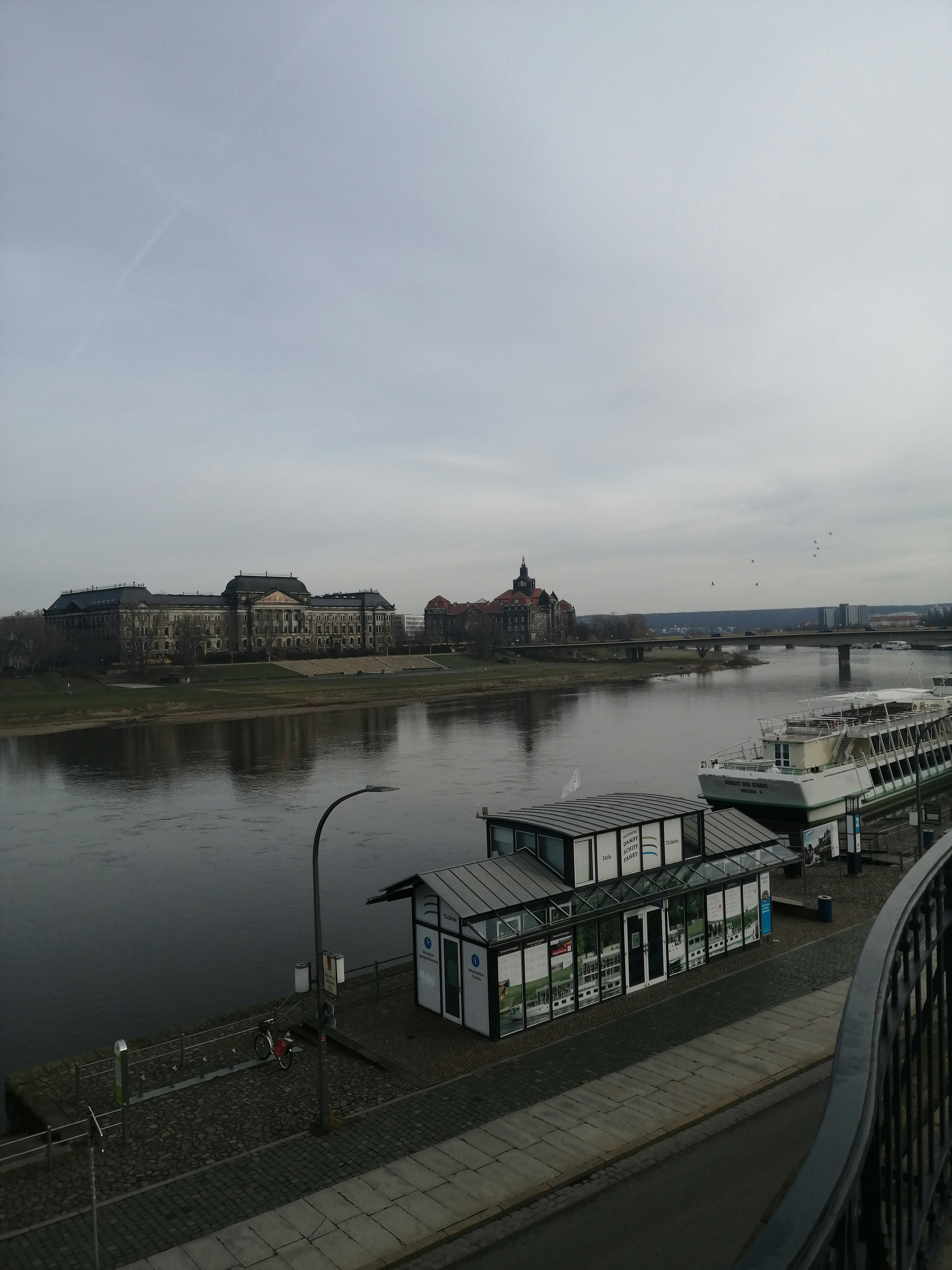 Riverside cityscape along a calm river with historic brick buildings and a prominent red-domed tower against a pale sky.
