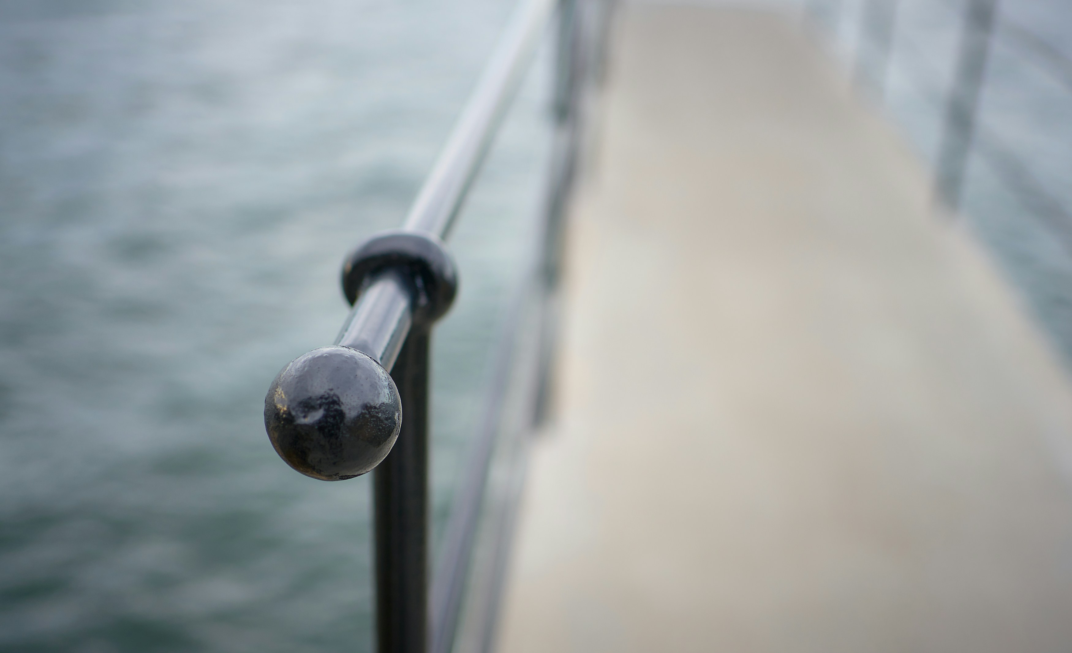 Close-up of a black railing on a pier, leading towards calm waters. The focus highlights the texture and details of the railing against a blurred background.