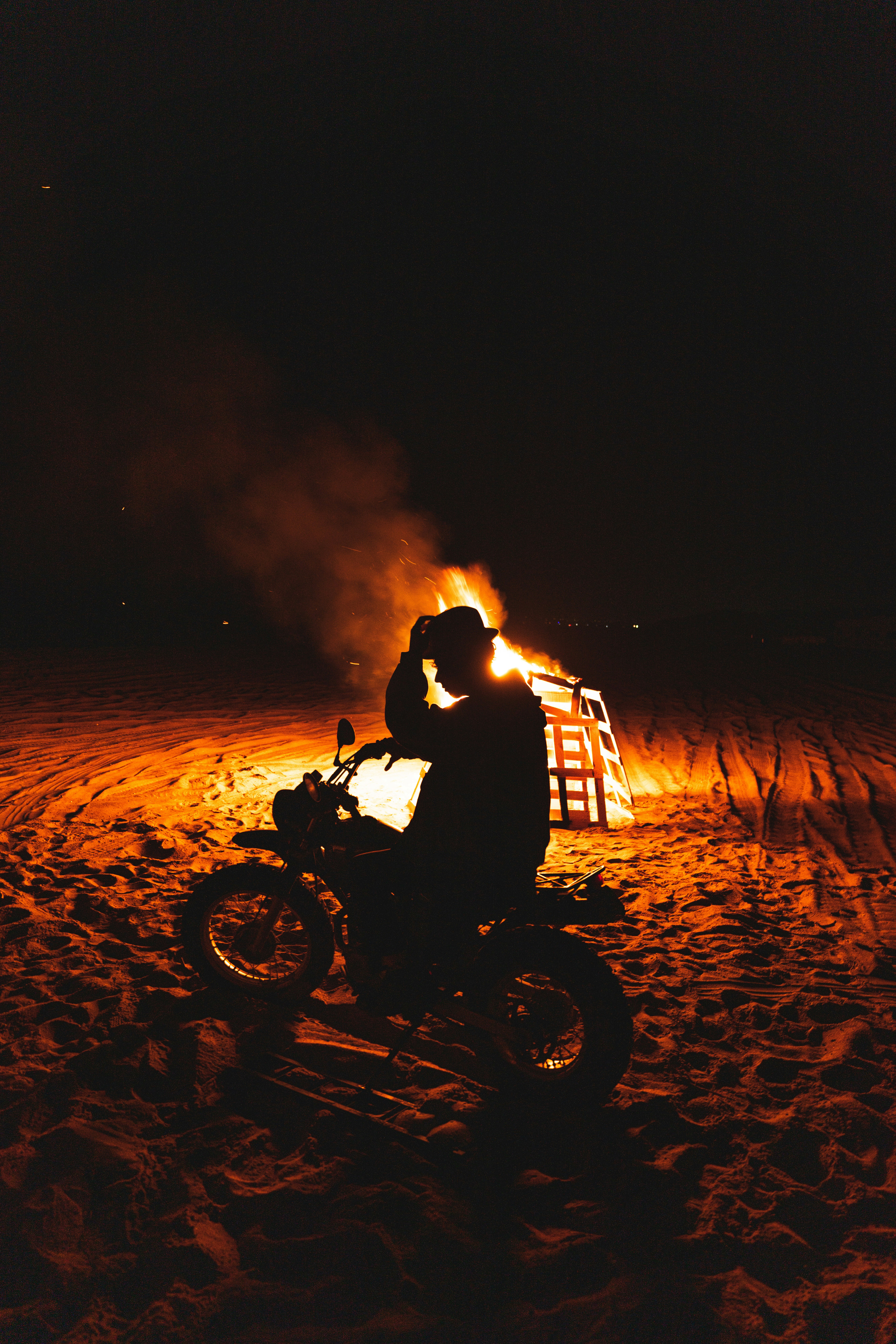 Silhouette of a rider on a motorcycle, capturing a moment of contemplation beside a flickering bonfire on a sandy beach.