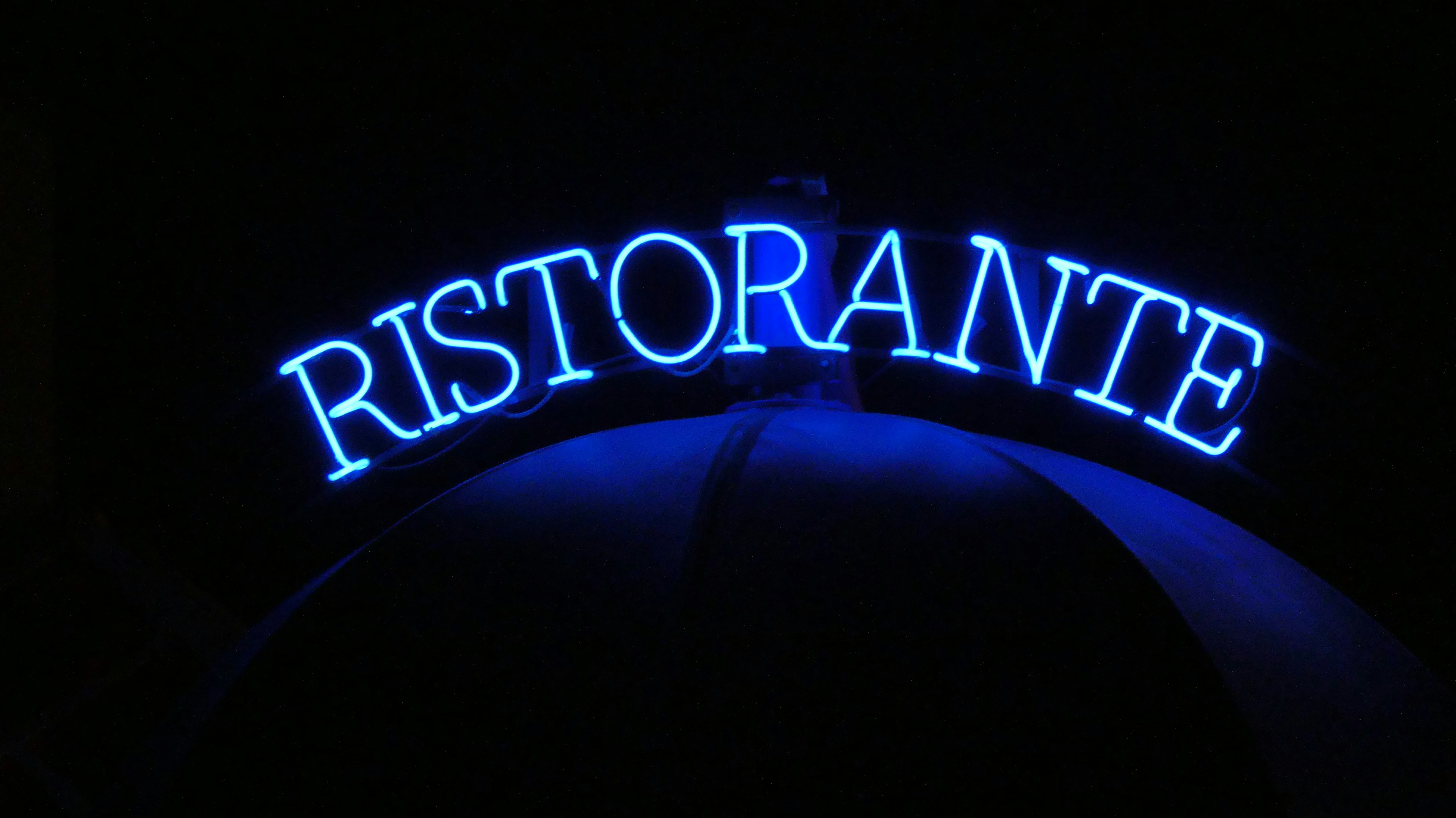 Blue neon RISTORANTE sign arches over a dark street, casting a cool glow on a curved canopy.