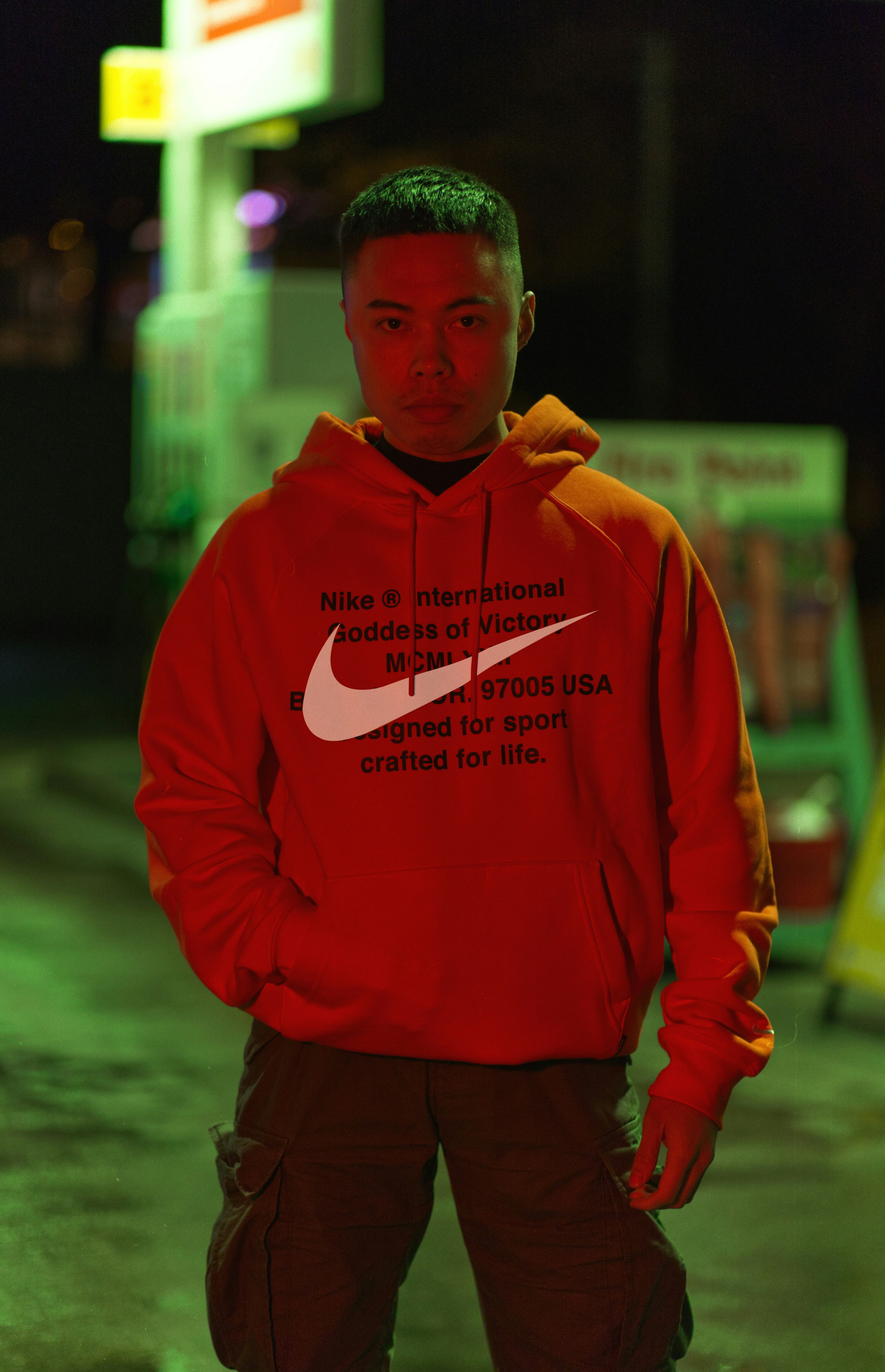 A young man stands confidently in an orange hoodie, illuminated by vibrant neon lights in an urban setting.