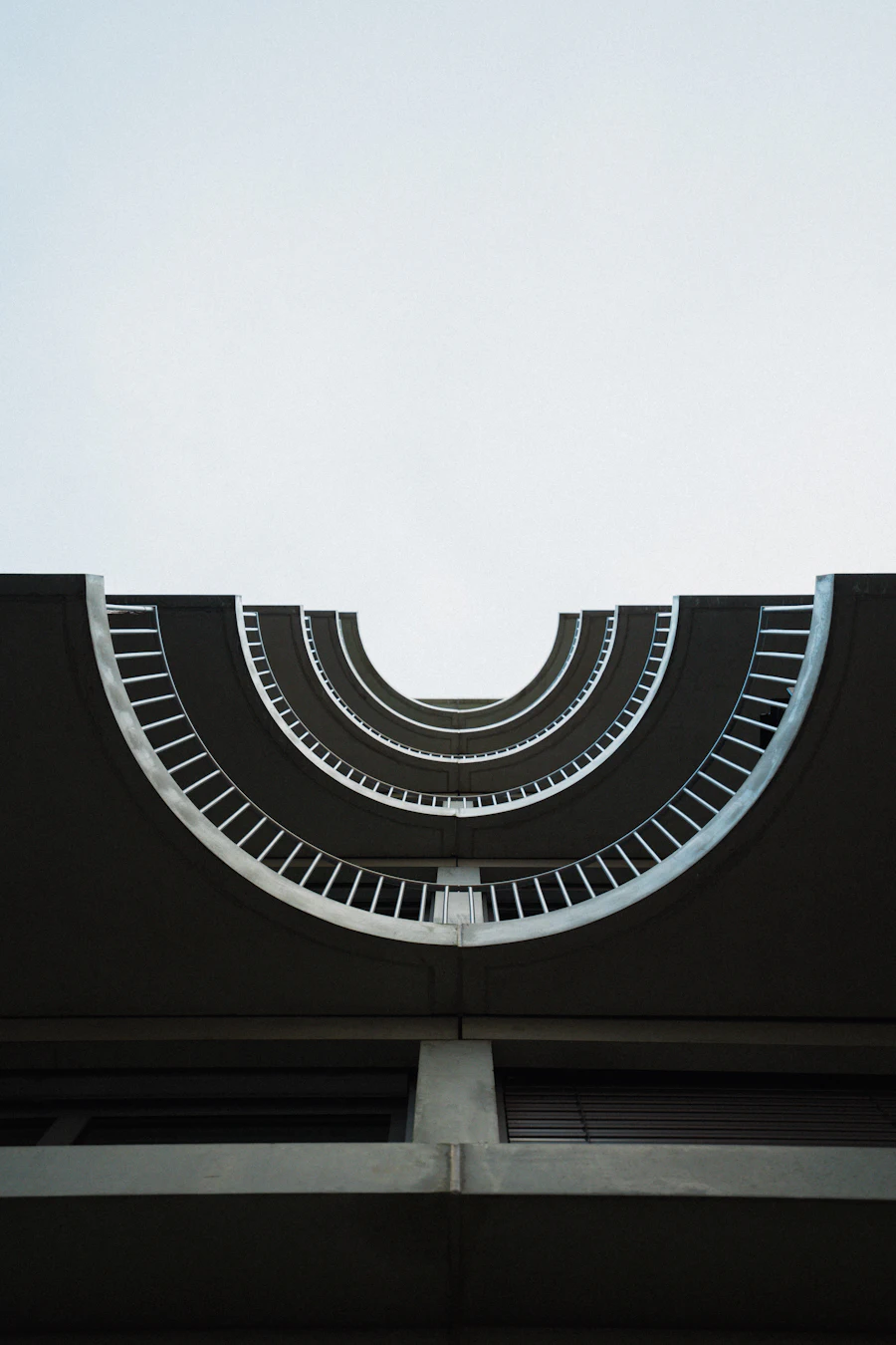 Dark modern building facade with repeating semicircular balconies against a pale sky. The architectural lines create a strong symmetrical pattern.