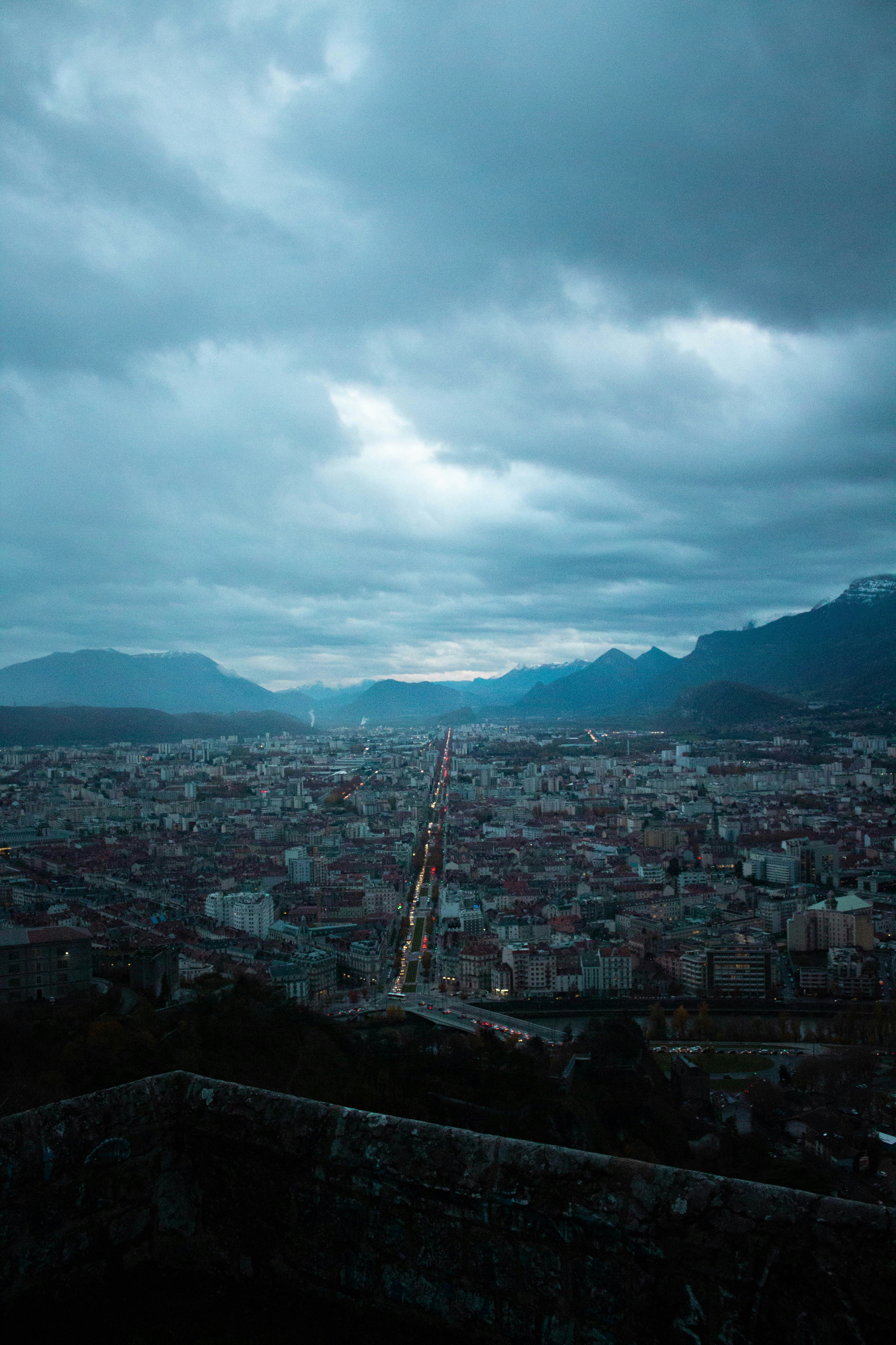 A panoramic view of a cityscape at dusk, showcasing a central avenue illuminated by streetlights, framed by distant mountains and a moody sky.