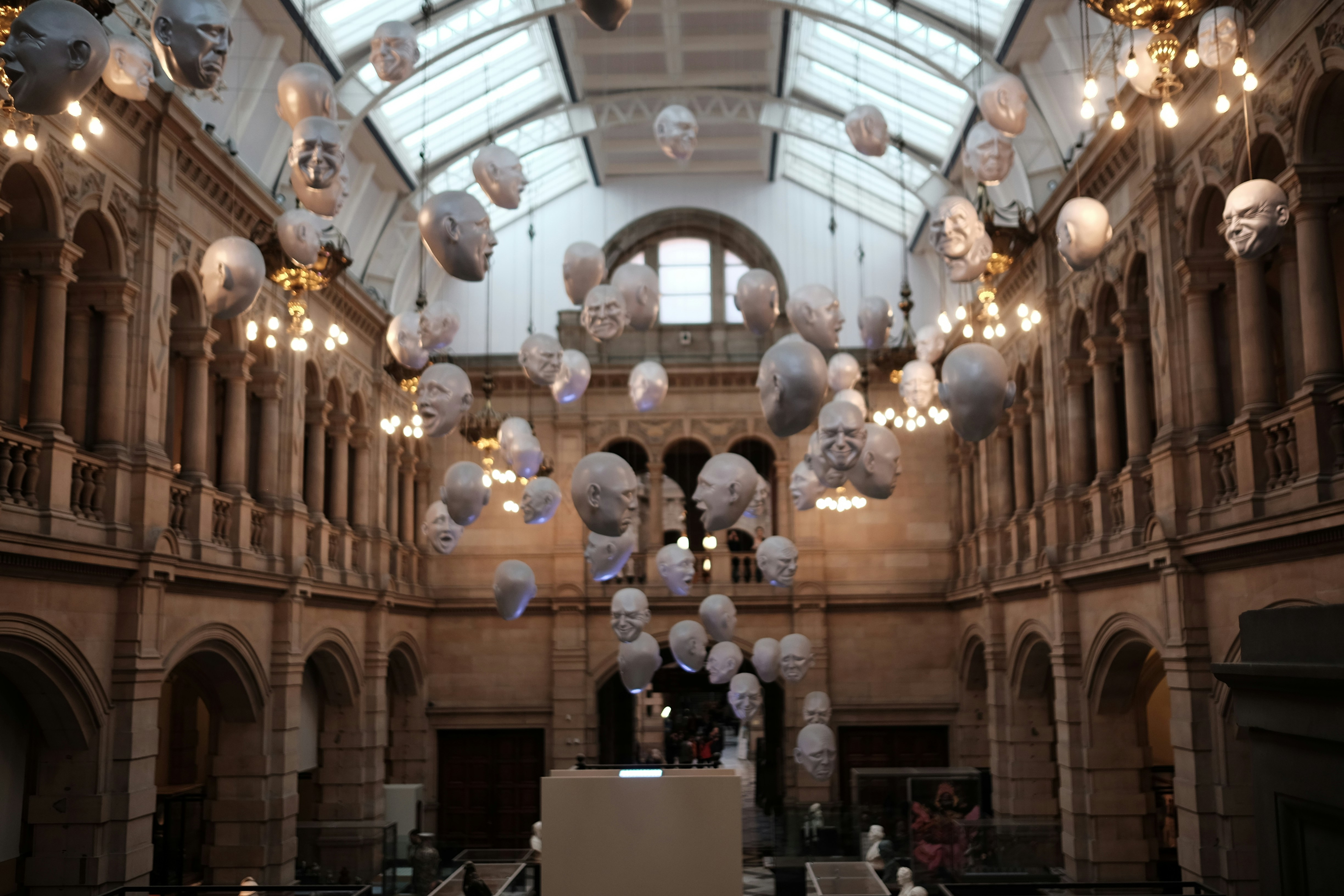 Sculptural heads suspended in a grand hall with arched windows and chandeliers.