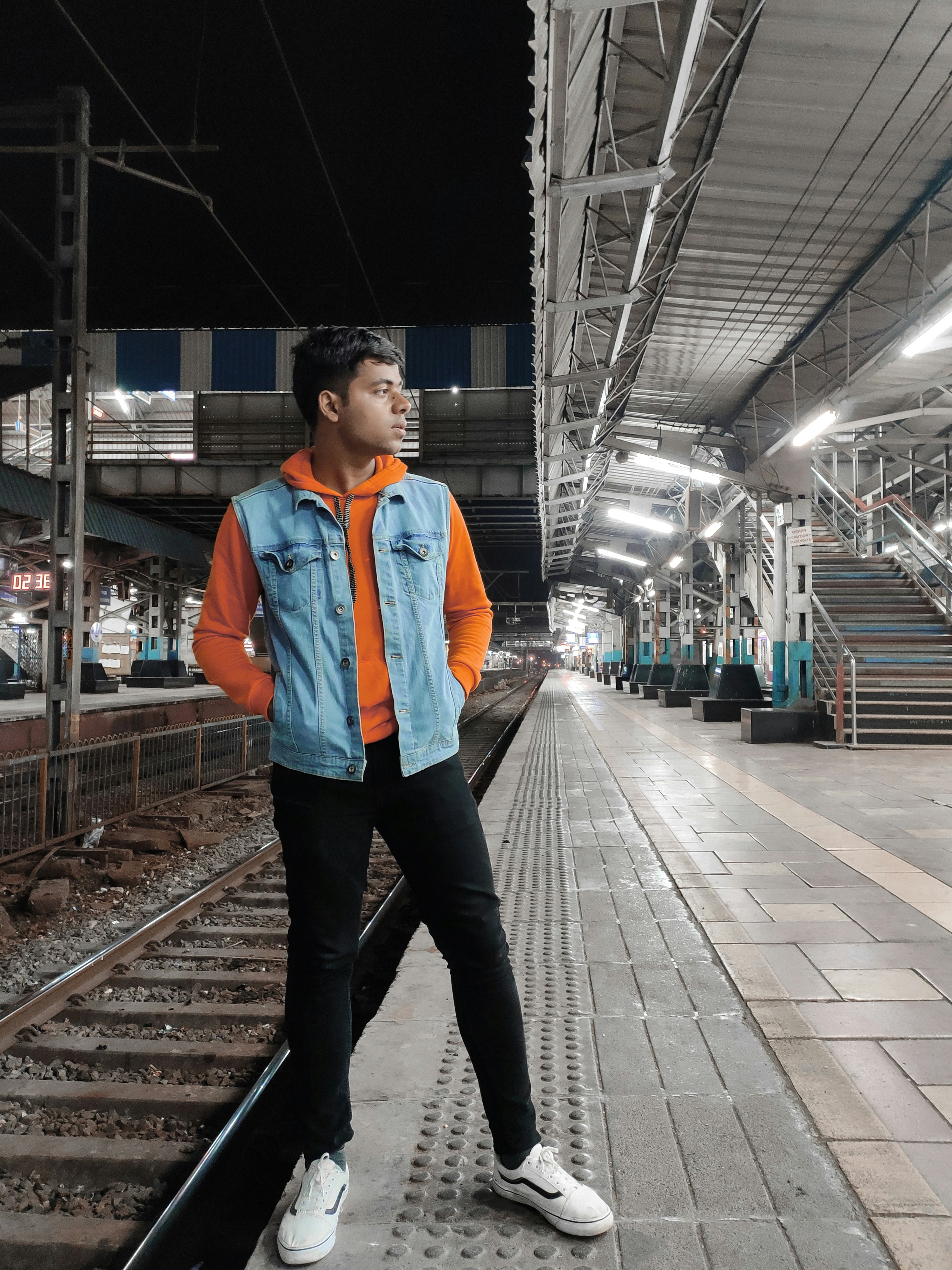 A young man stands thoughtfully at a train station platform, surrounded by industrial architecture and dim lighting. The scene captures a blend of modern fashion and urban ambiance.