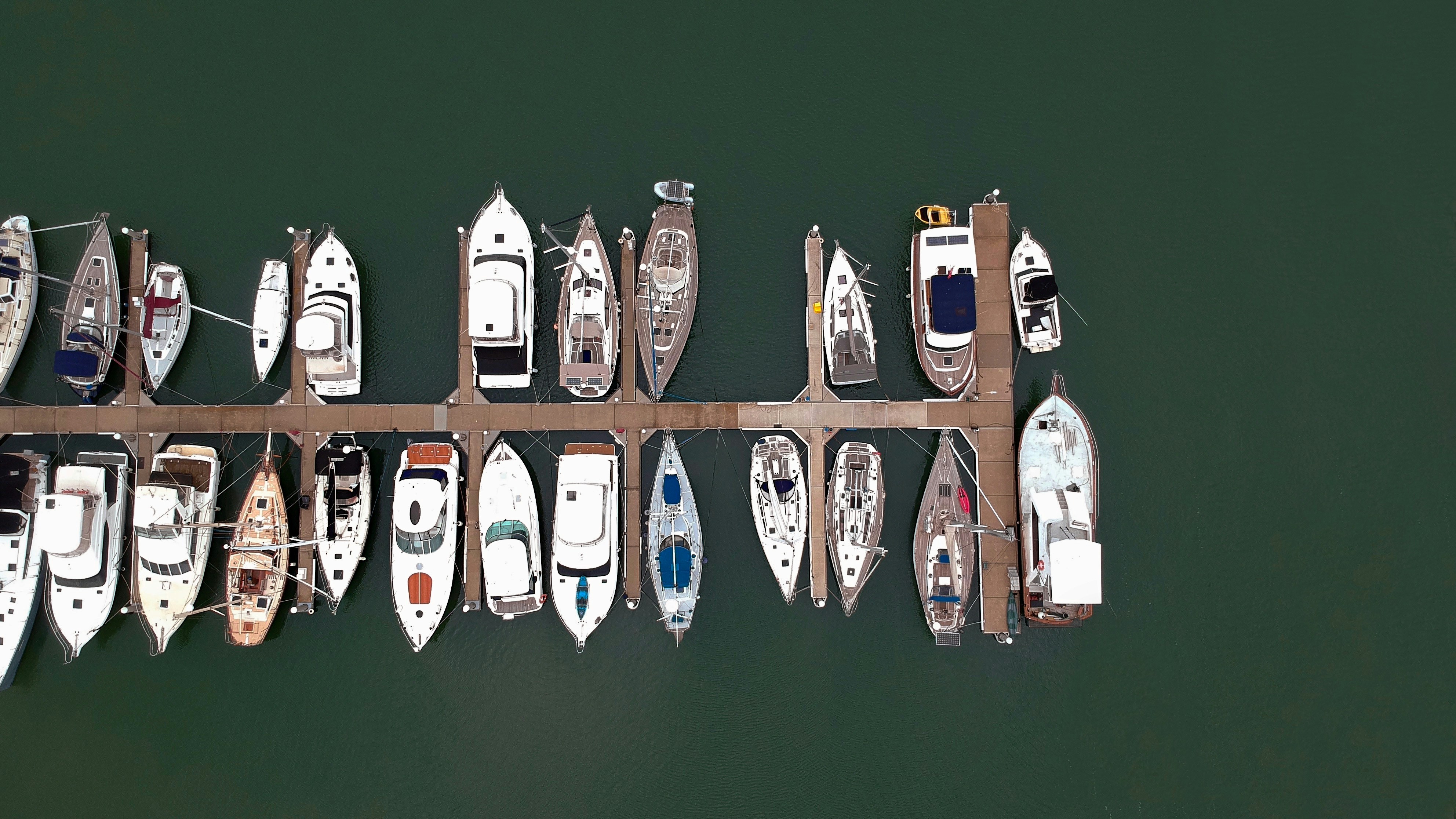 Aerial view of various boats neatly arranged at a marina, showcasing their diverse designs and colors against a dark green water backdrop.