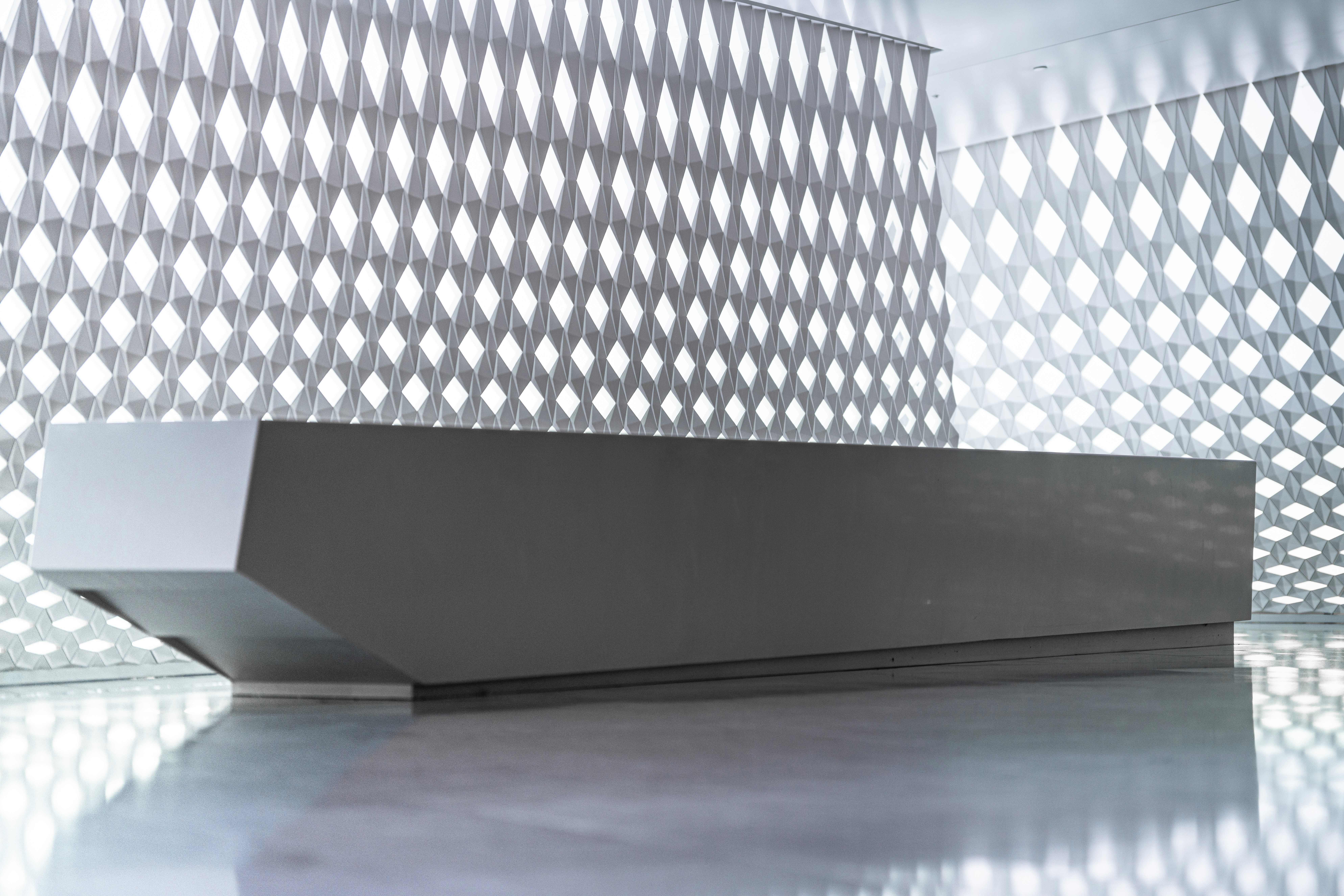 A photorealistic interior photograph featuring a grey, modular bench on a glossy floor. Diamond-pattern metal walls provide a futuristic backdrop that emphasizes the bench's sculptural geometry.