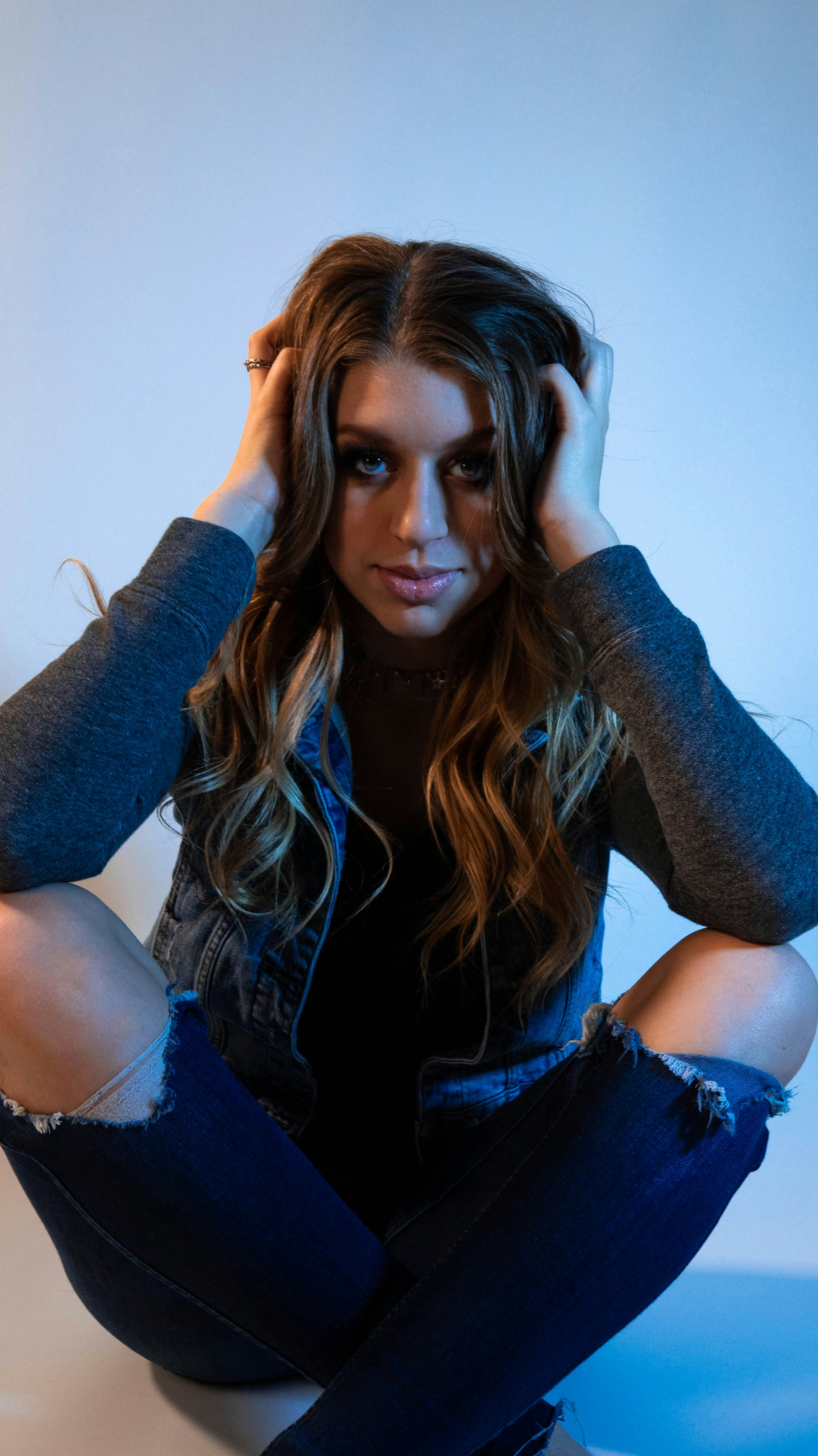 Portrait of a woman seated with knees drawn up, wearing a denim jacket and hoodie under cool blue studio lighting. The subdued setup emphasizes her focused gaze.