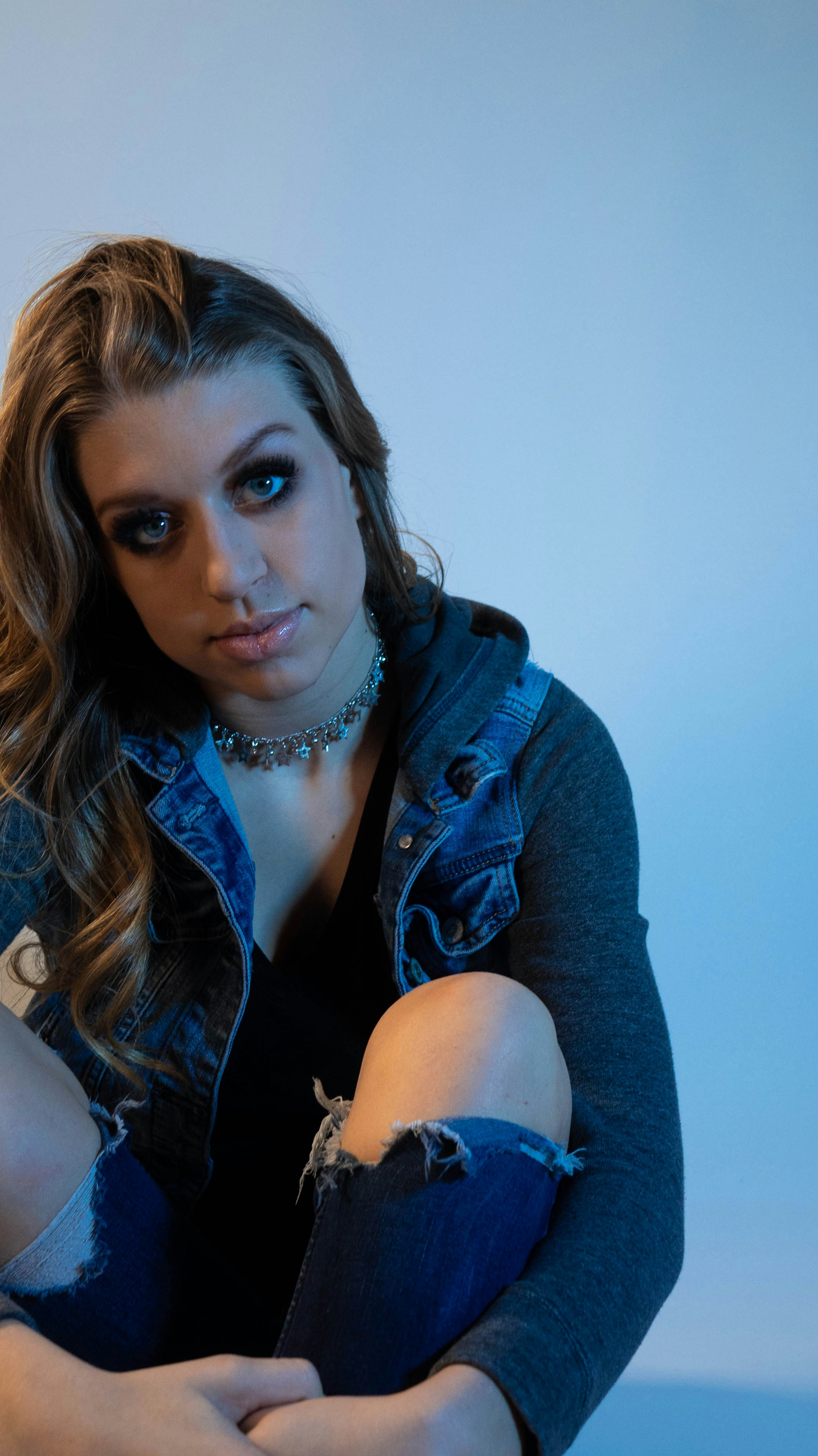 Portrait of a woman in a denim jacket under blue studio lighting, with a moody gaze. The photograph emphasizes color, texture, and the subject's expression against a minimal backdrop.