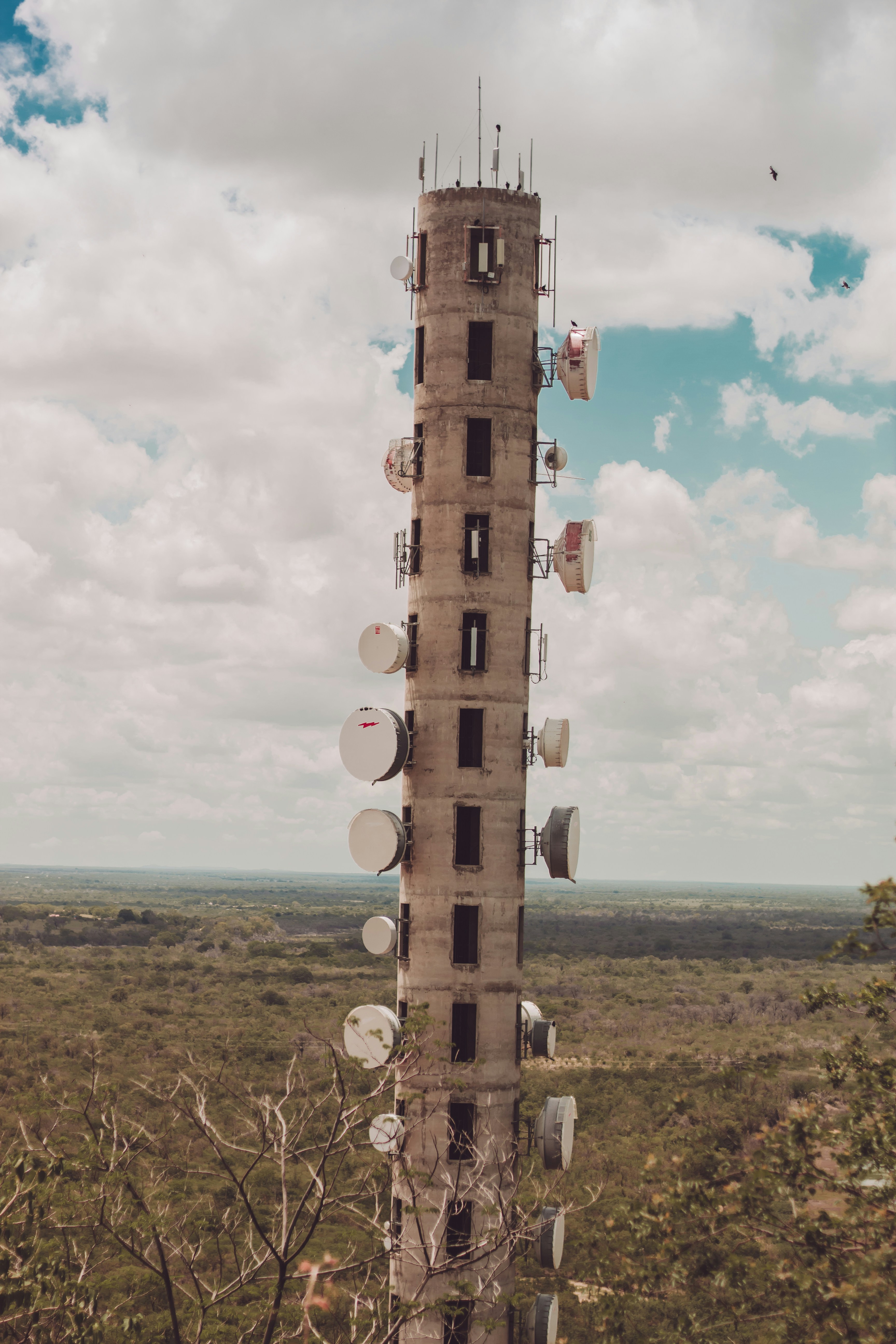 A tall telecommunications tower adorned with satellite dishes rises above a lush, green landscape under a partly cloudy sky.