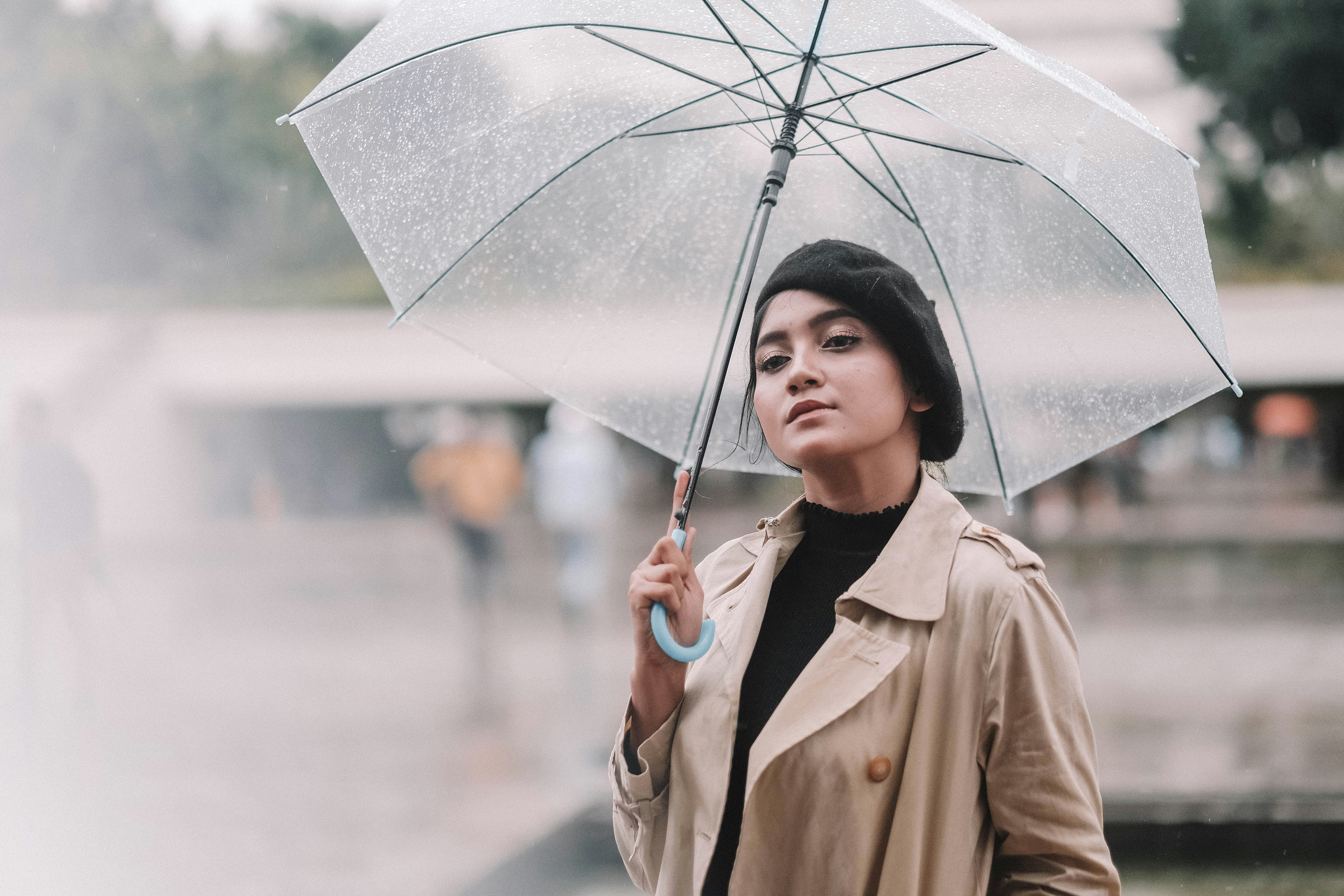A woman in a trench coat holds a clear umbrella, gazing thoughtfully amidst a misty urban backdrop. The scene captures the essence of a rainy day.