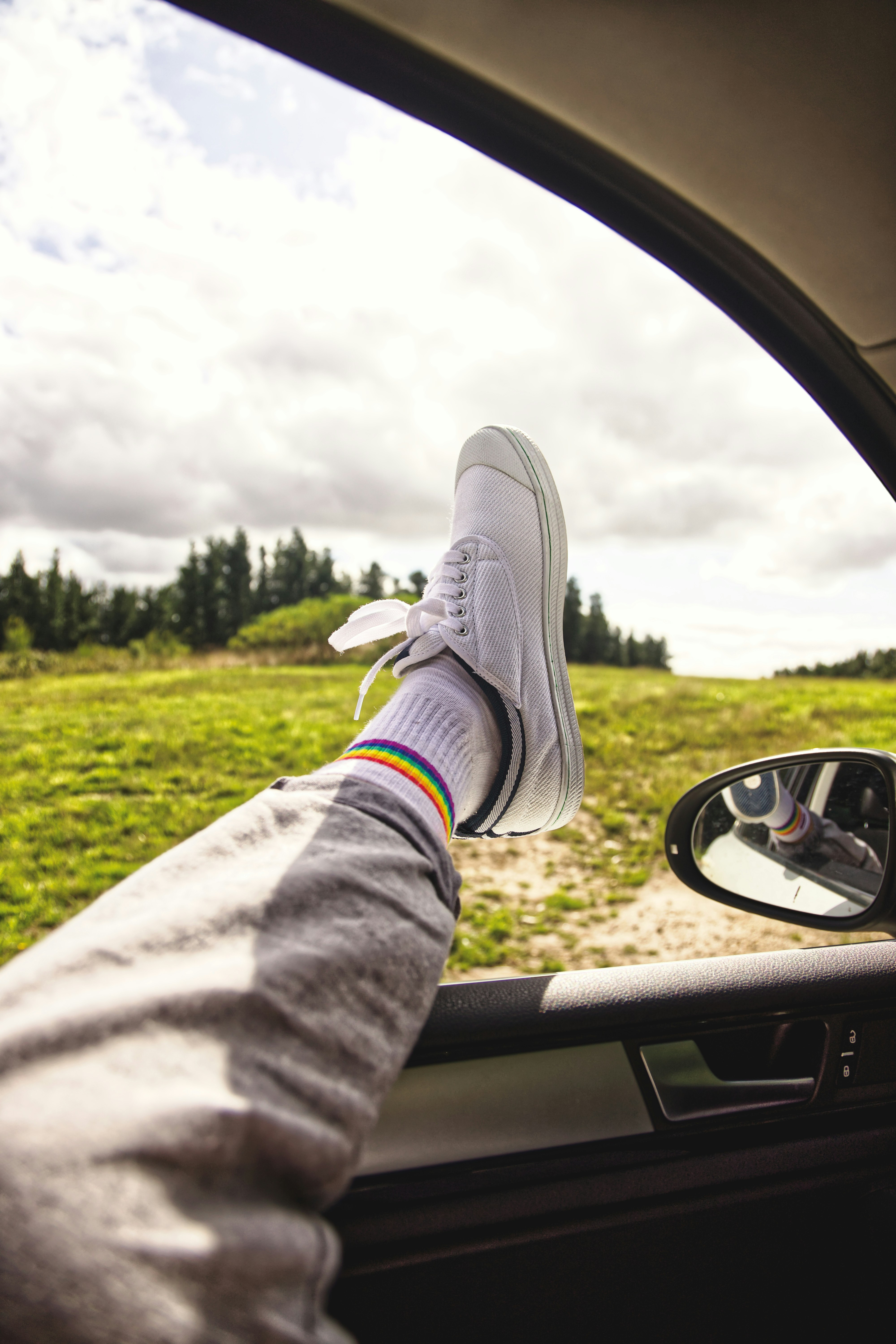 Window-side Escape: White sneaker with rainbow socks resting on an open car frame against a sunlit field.