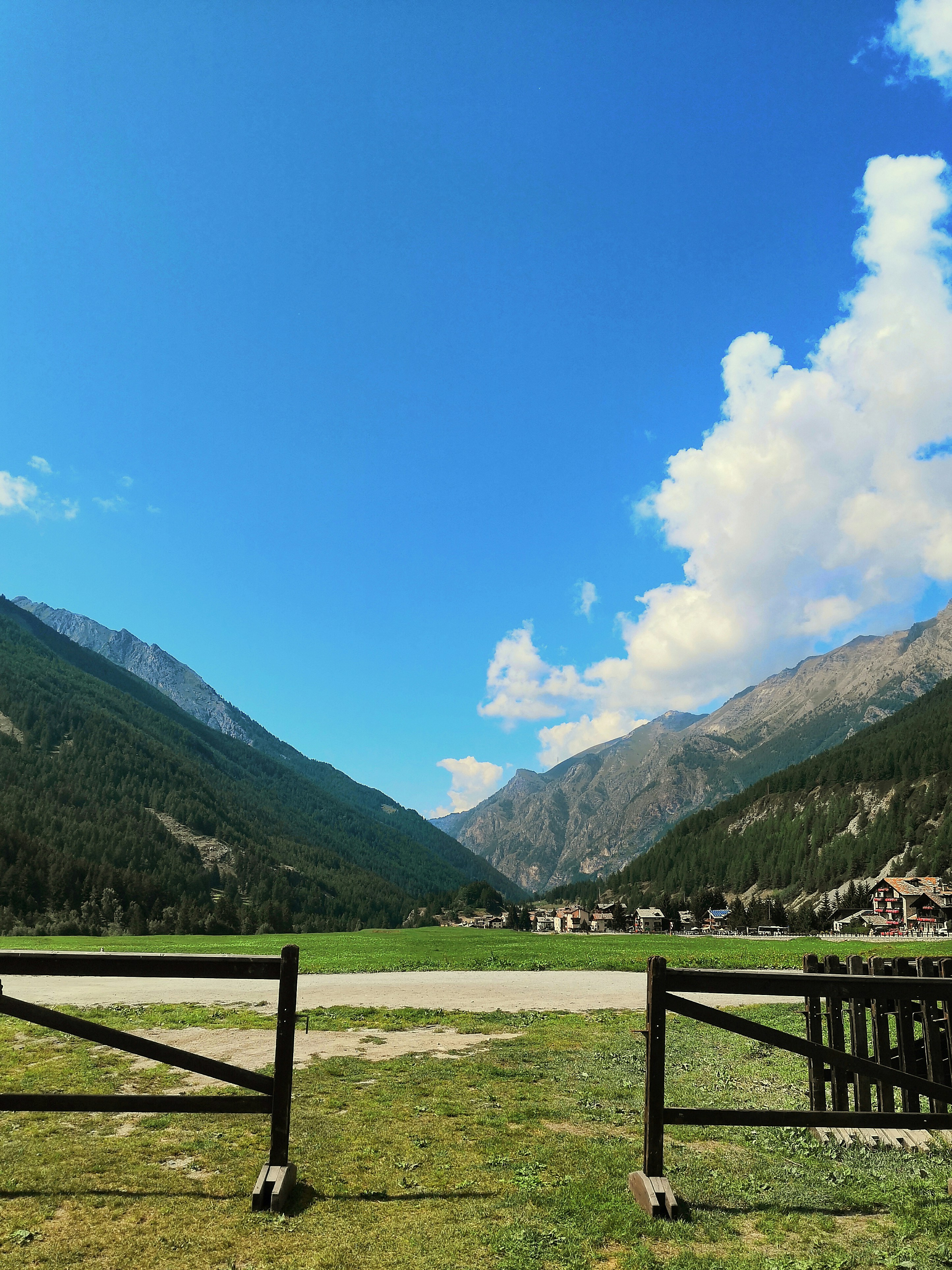 Expansive valley framed by majestic mountains under a vibrant blue sky with scattered clouds. A rustic wooden fence adds depth to the serene landscape.