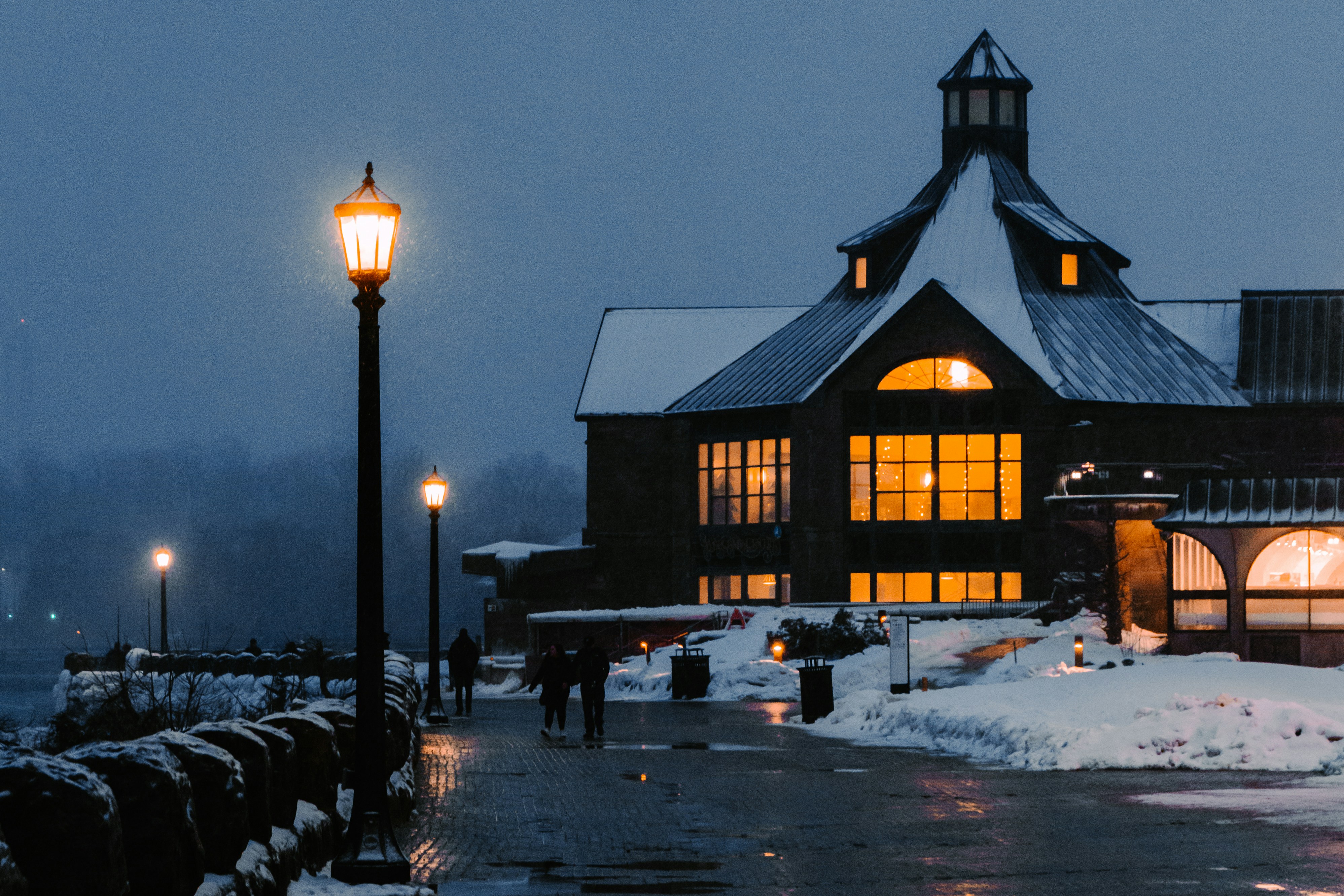 Cozy building illuminated by warm lights amidst a snowy landscape at dusk, with lampposts lining the path. 