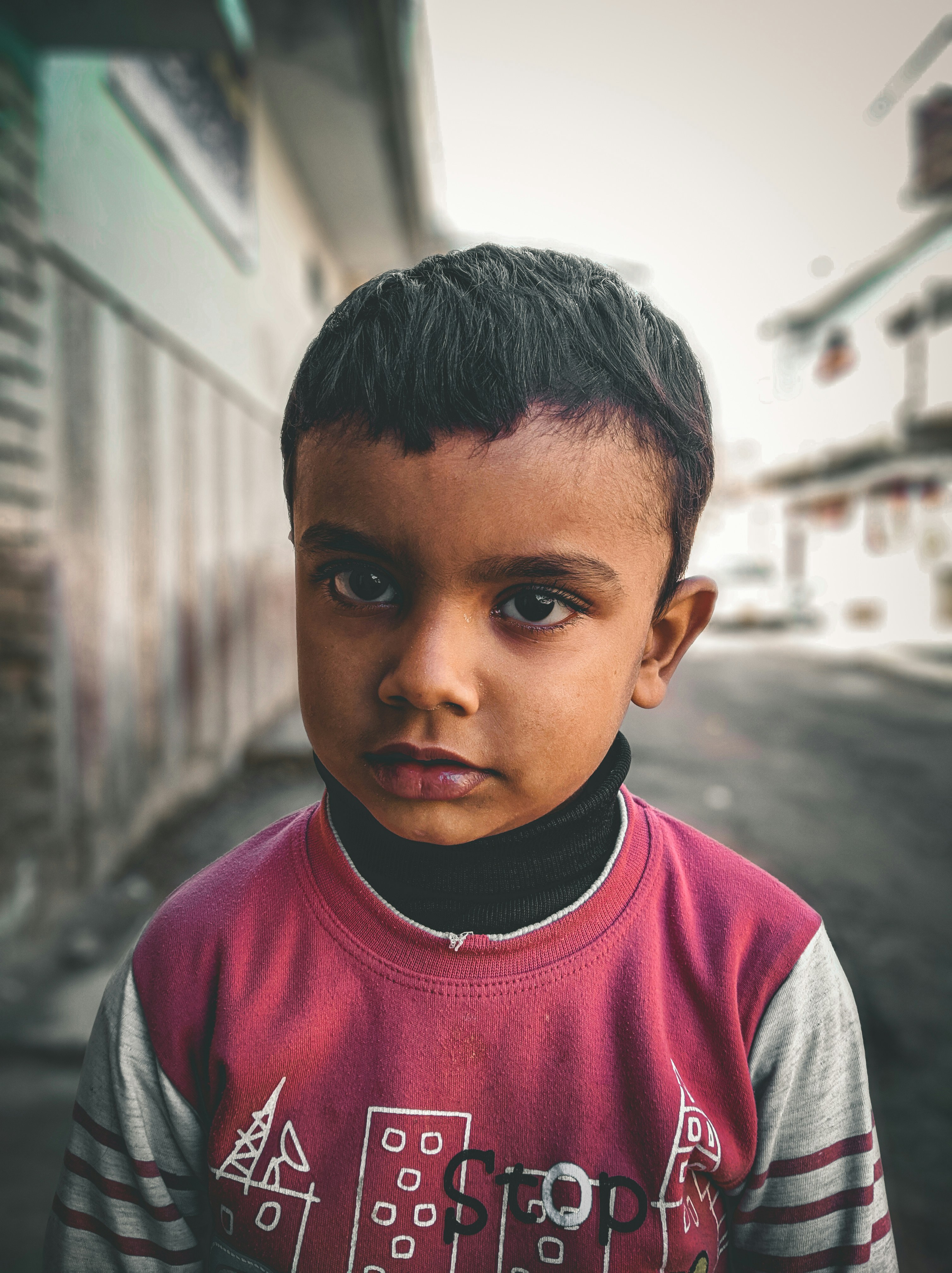 Young boy with a serious expression stands against a blurred urban backdrop, highlighting his contemplative demeanor.