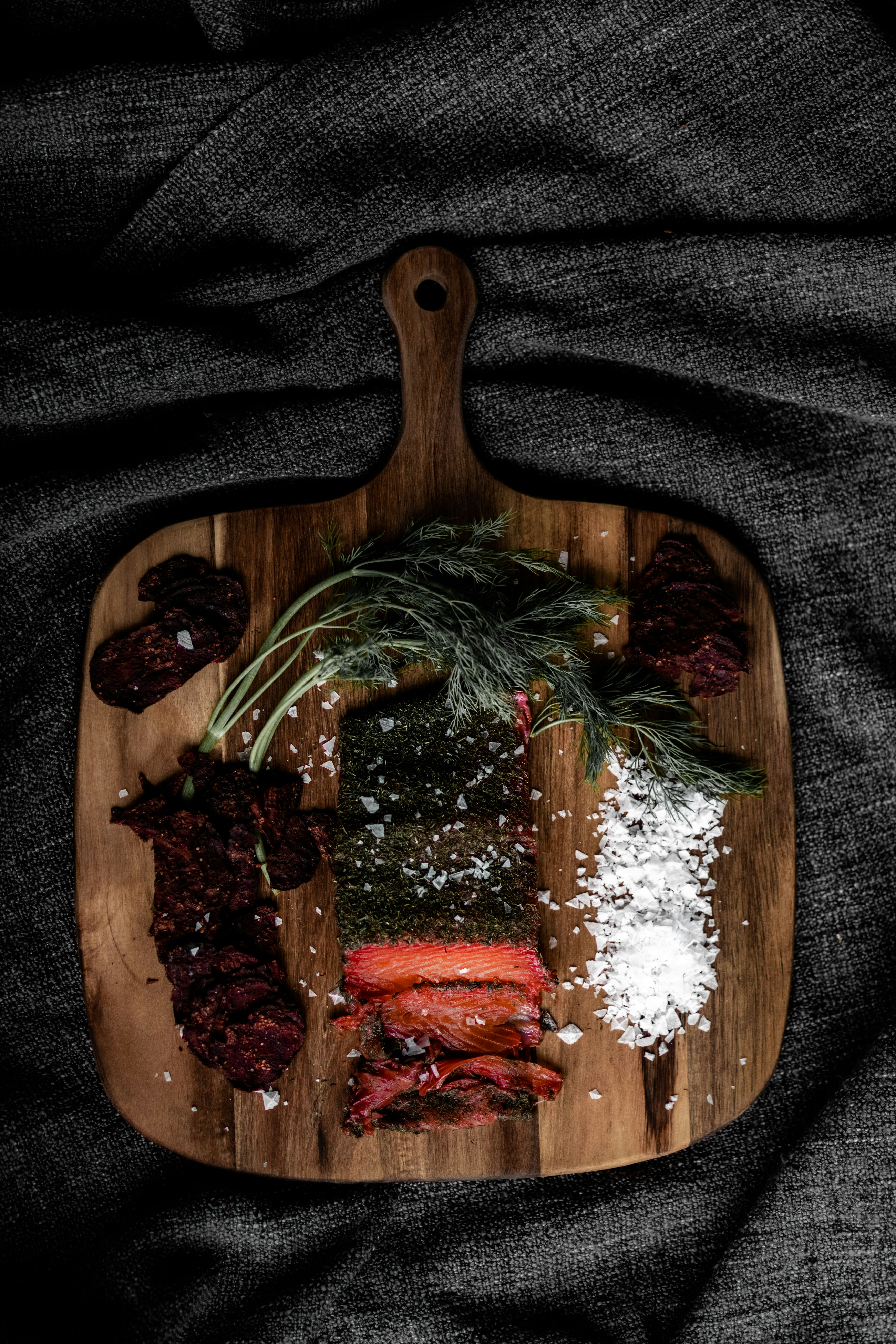 A wooden cutting board displays an artistic arrangement of smoked salmon, herbs, and garnishes, highlighting a culinary masterpiece. Flaky sea salt adds a touch of elegance.