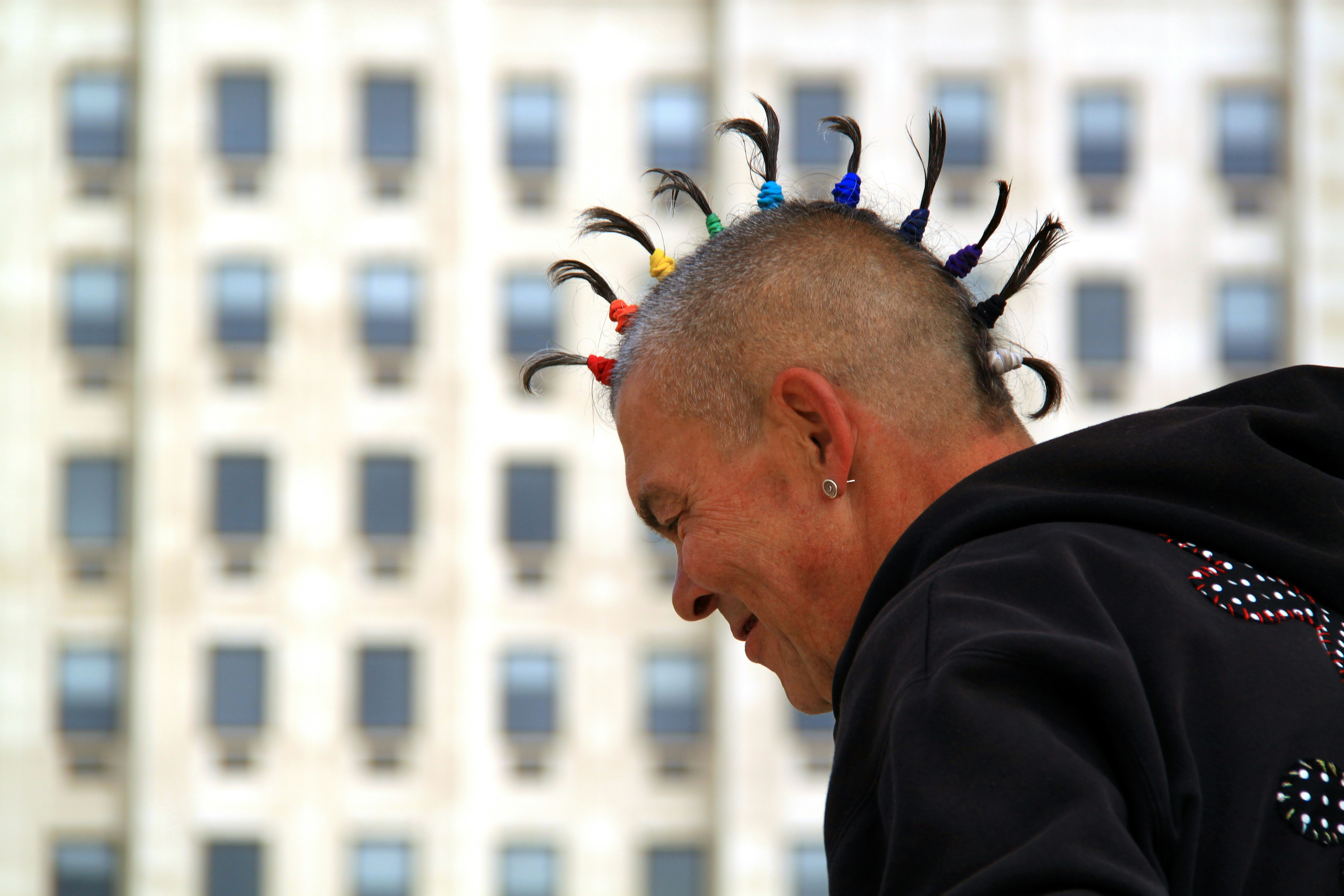 Man with rainbow-colored mohawk against a blurred urban building backdrop.