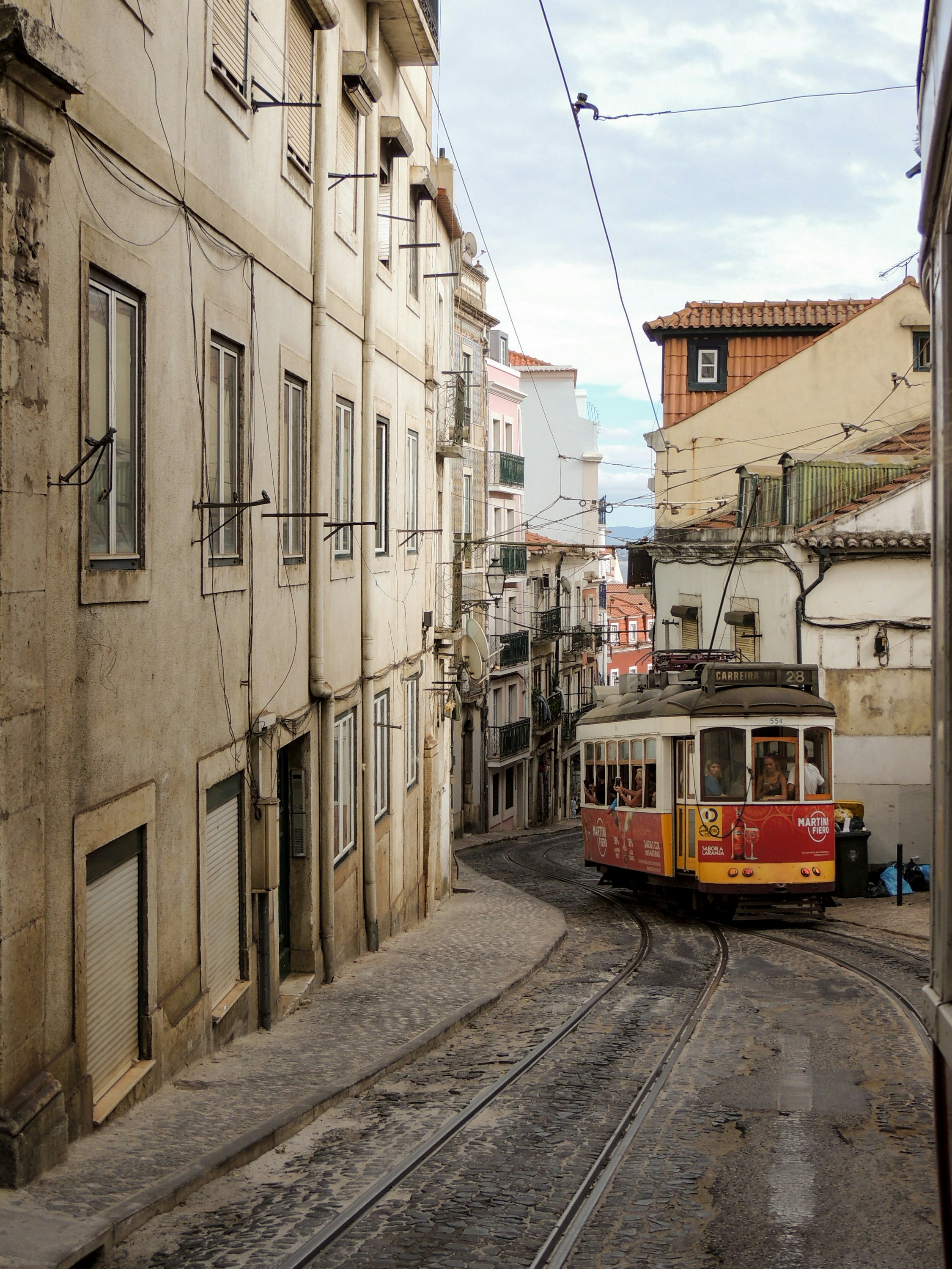 Red tram winds through a steep, narrow cobblestone street lined with aging pastel buildings, as overhead wires crisscross above.