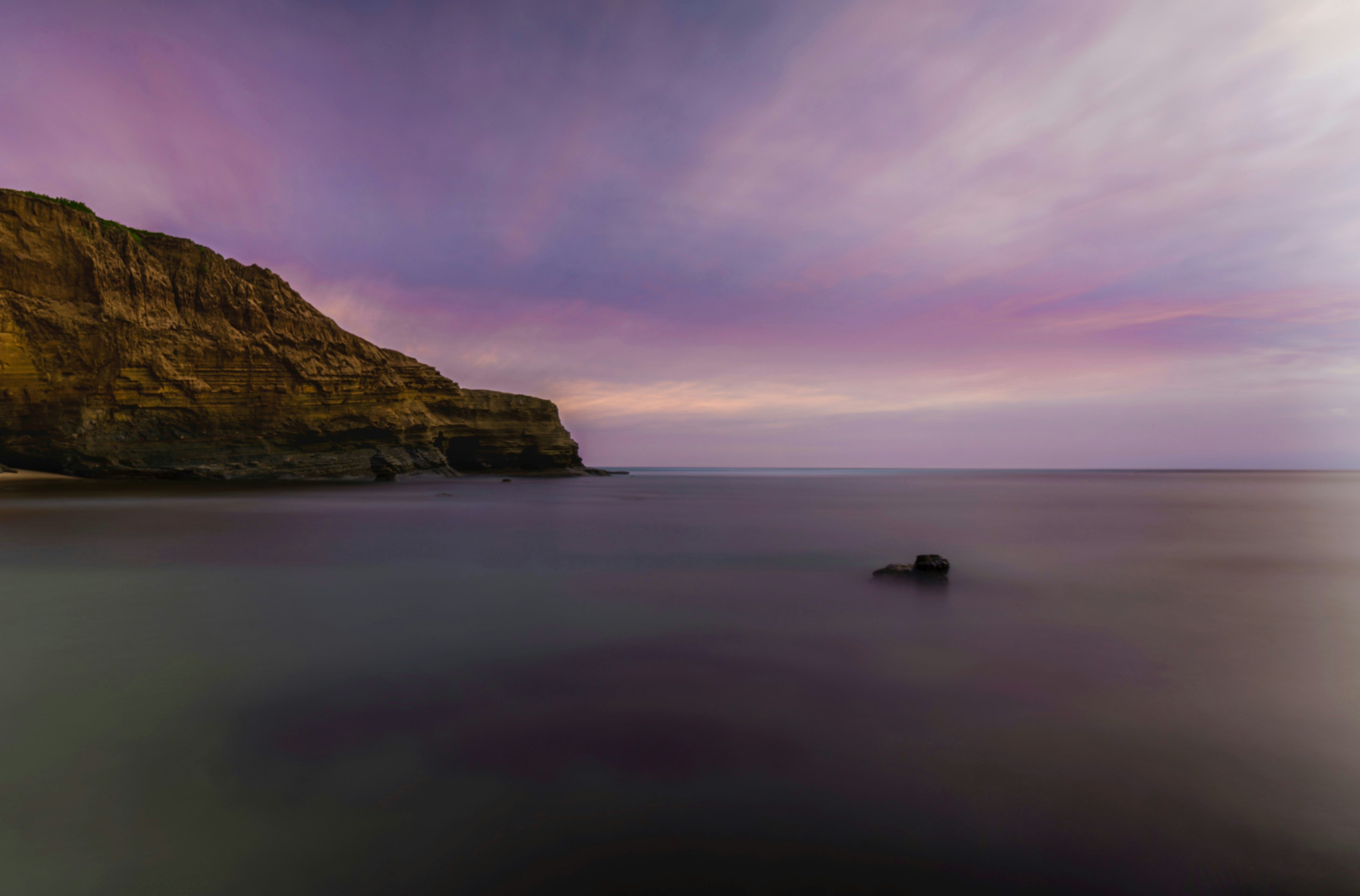 Golden cliffs silhouetted against a soft gradient of twilight hues, reflecting on calm waters. A solitary rock emerges from the serene surface.